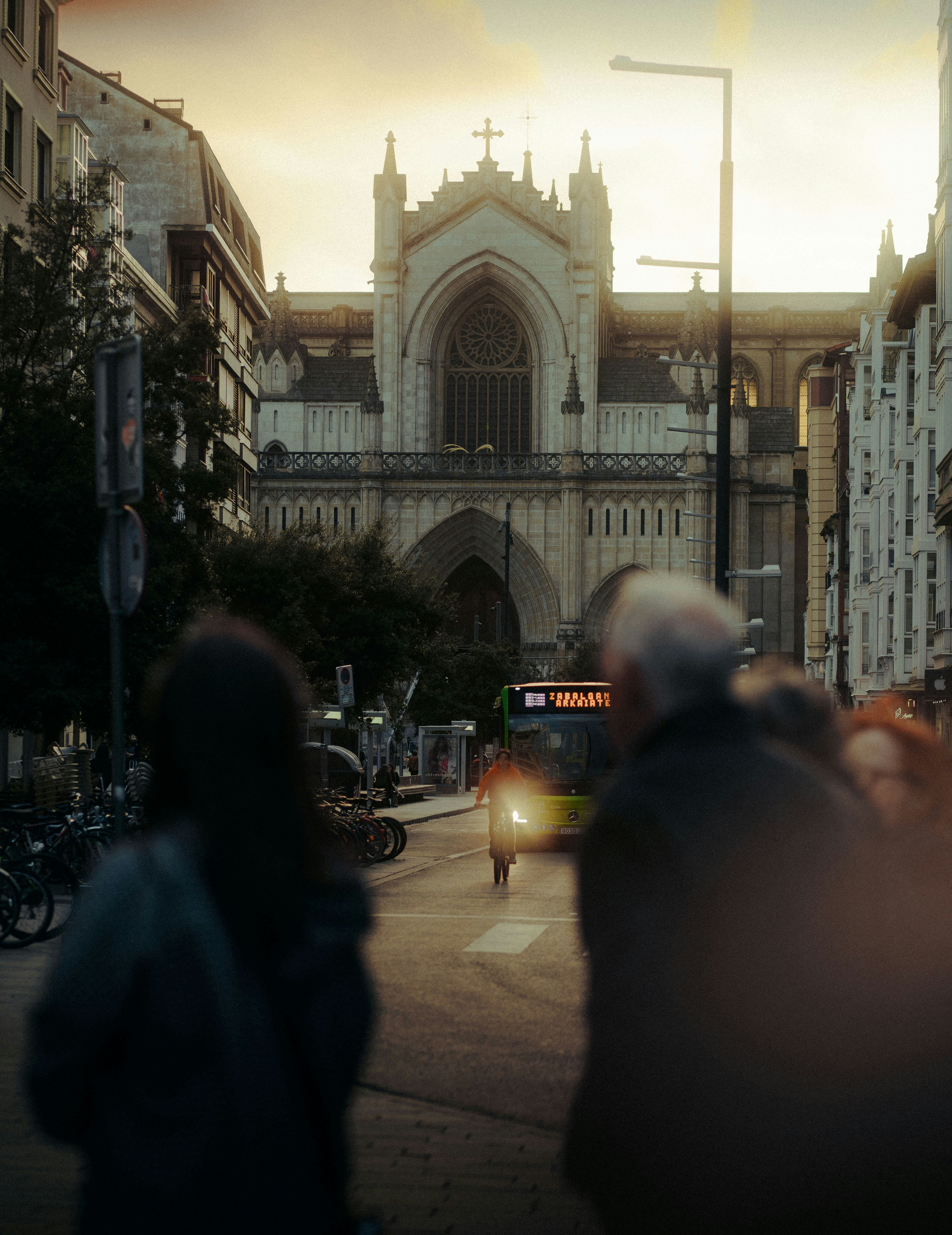 Gothic cathedral looms majestically over a bustling city street, framed by pedestrians and bicycles. The warm glow of sunset casts a serene ambiance.
