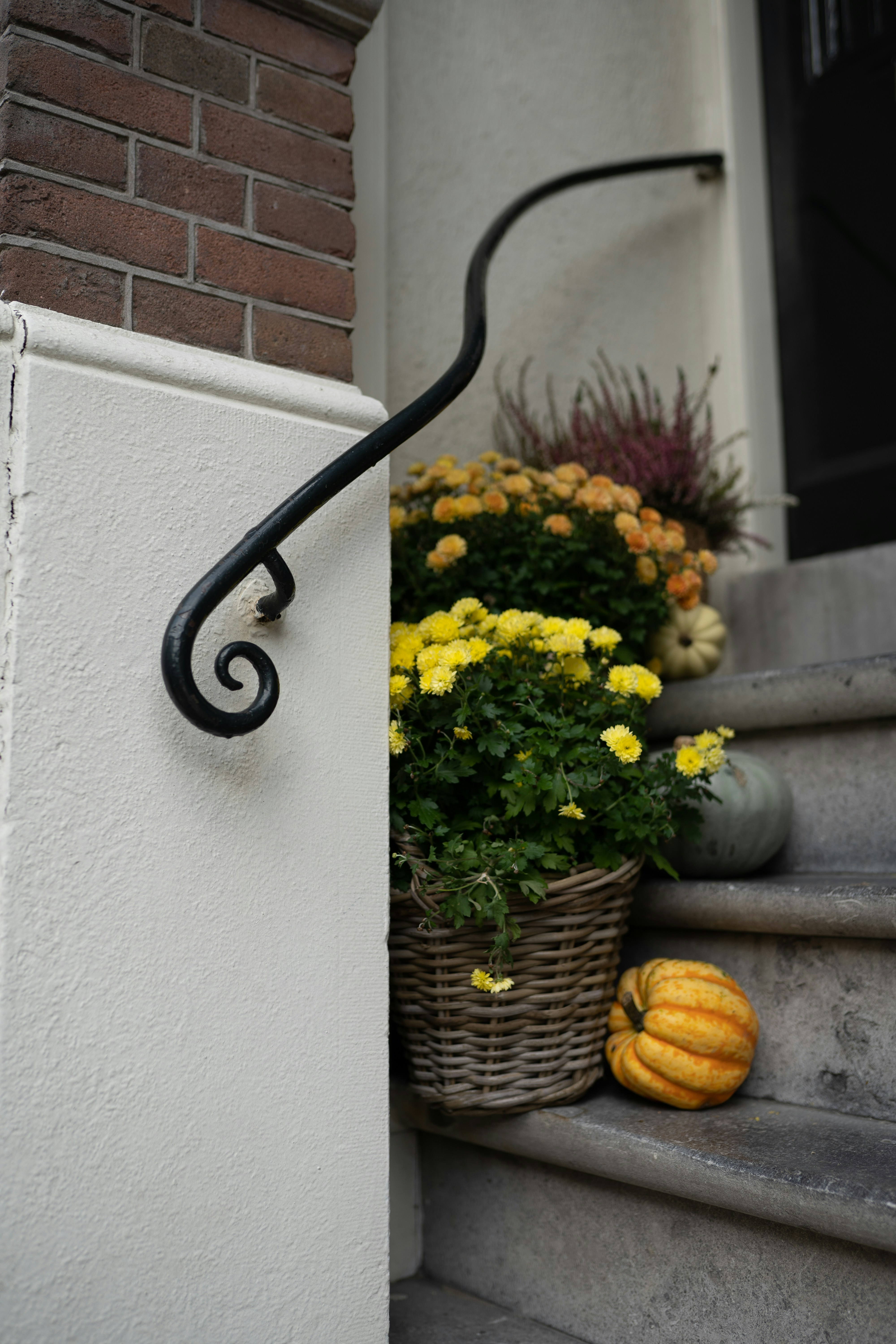Photograph of a wicker basket overflowing with yellow chrysanthemums beside stone steps, with small pumpkins and gourds nearby. A curved wrought-iron railing frames the scene.