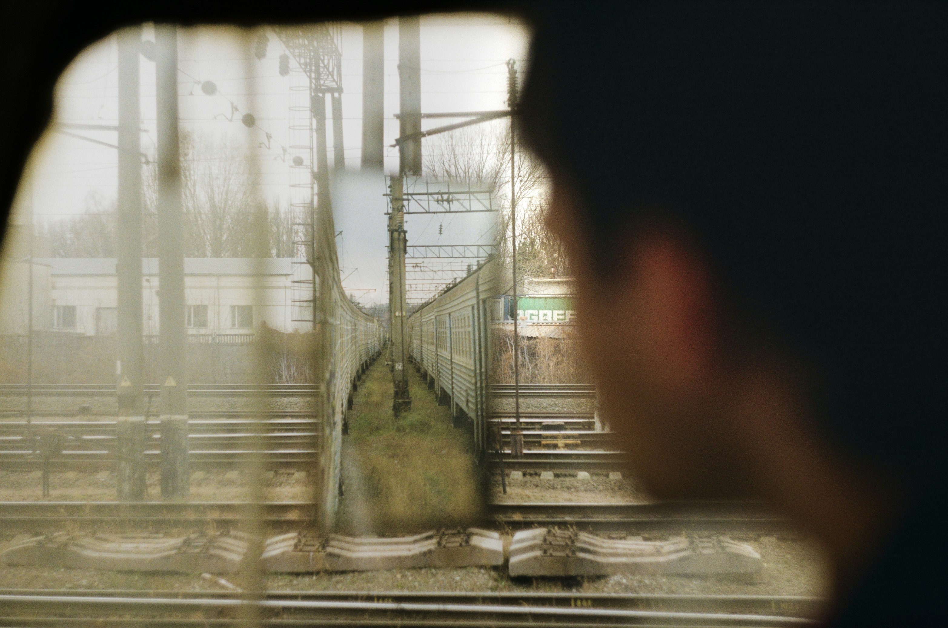 Rail tracks draw the eye toward a distant station framed by the view through a window. A blurred silhouette in the foreground adds depth and mystery.
