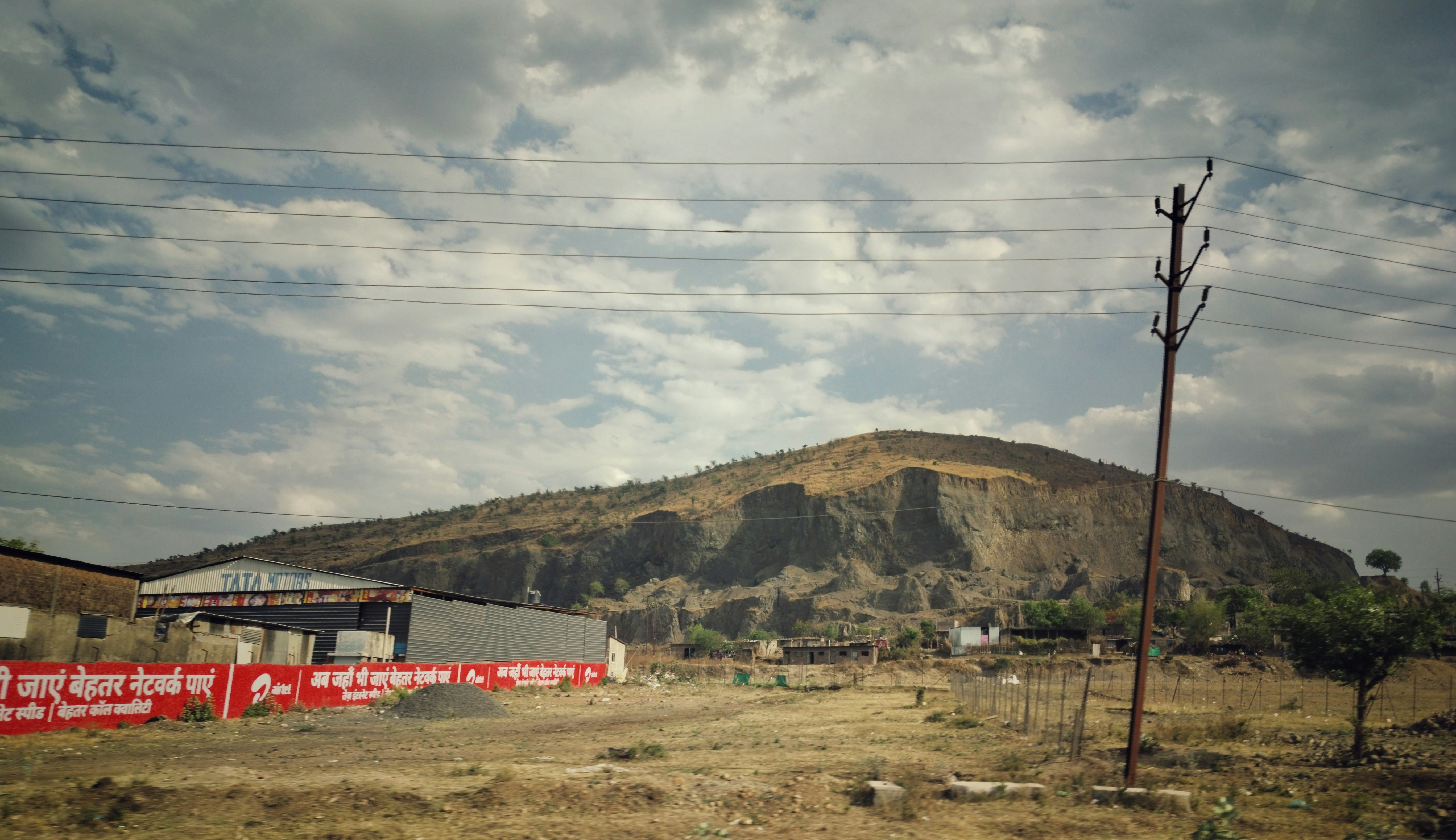 Rural landscape with a low hill, power lines, and a red-walled building under a cloudy sky.
