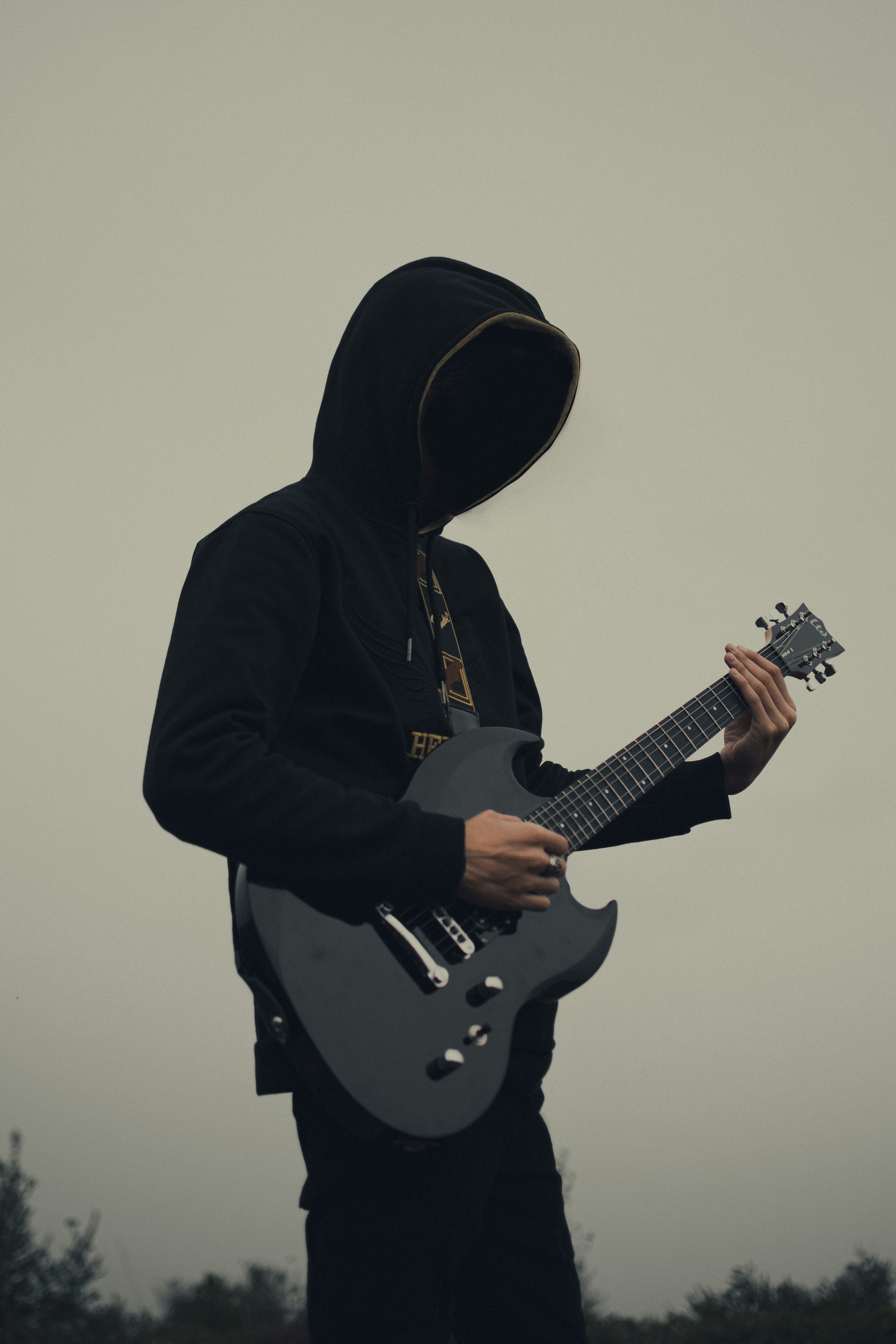 Hooded guitarist silhouette under a pale, desaturated sky with a matte electric guitar