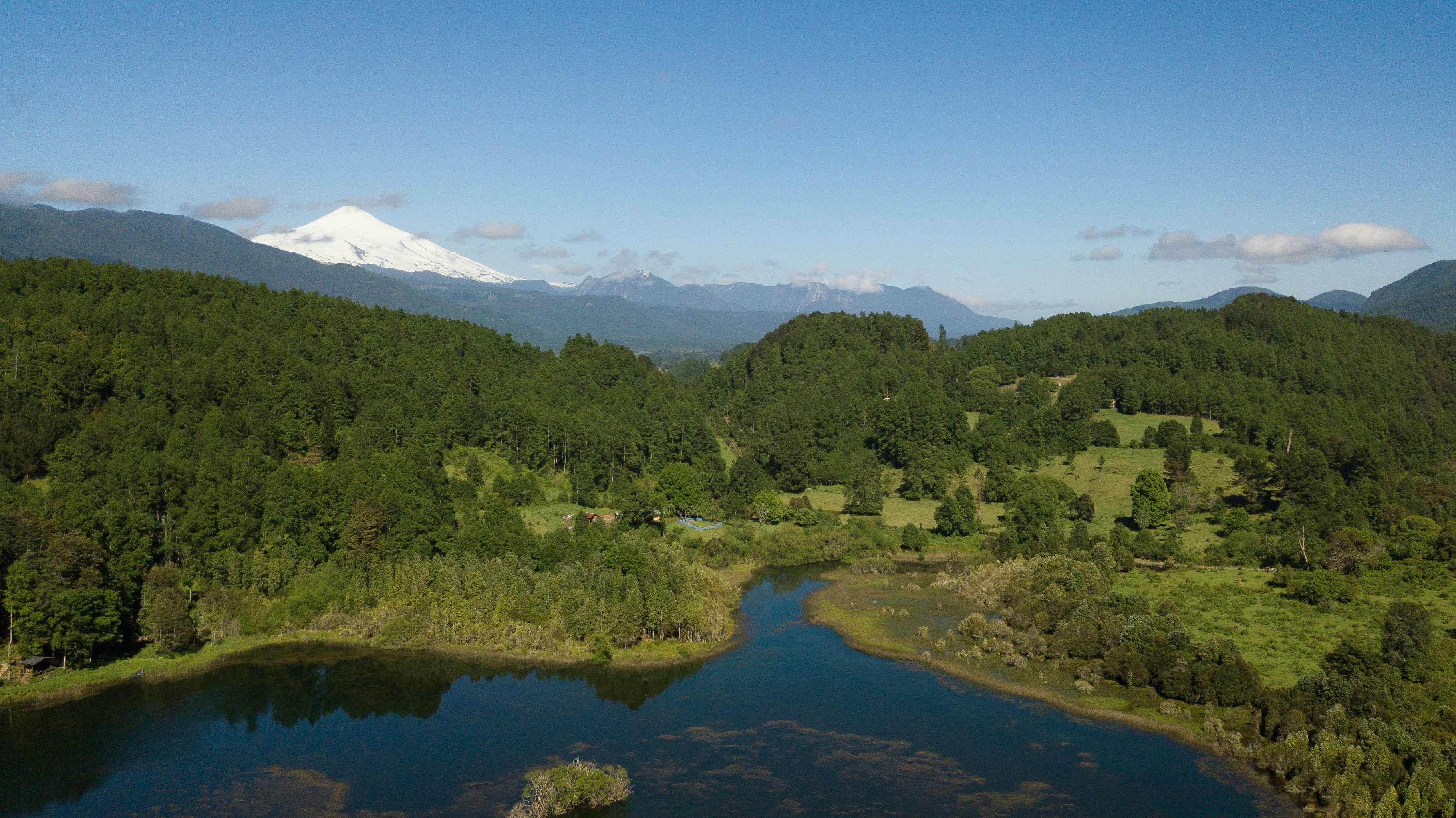 Lush green landscape with a river winding through forested hills under a clear blue sky, with a snow-capped mountain in the distance.