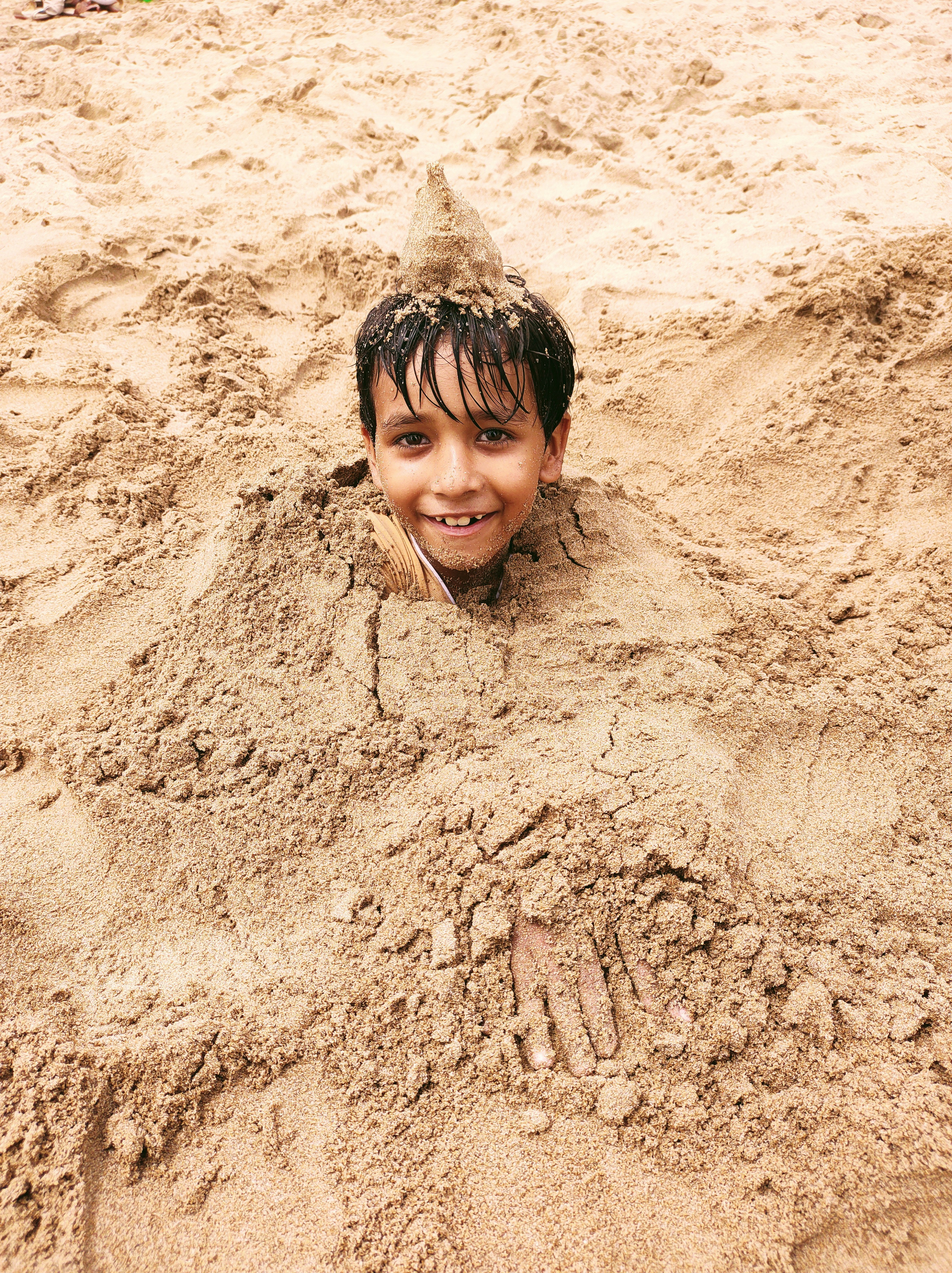 Photograph of a smiling boy peeking from a mound of sand on a sunny beach, with only his head and hands visible.