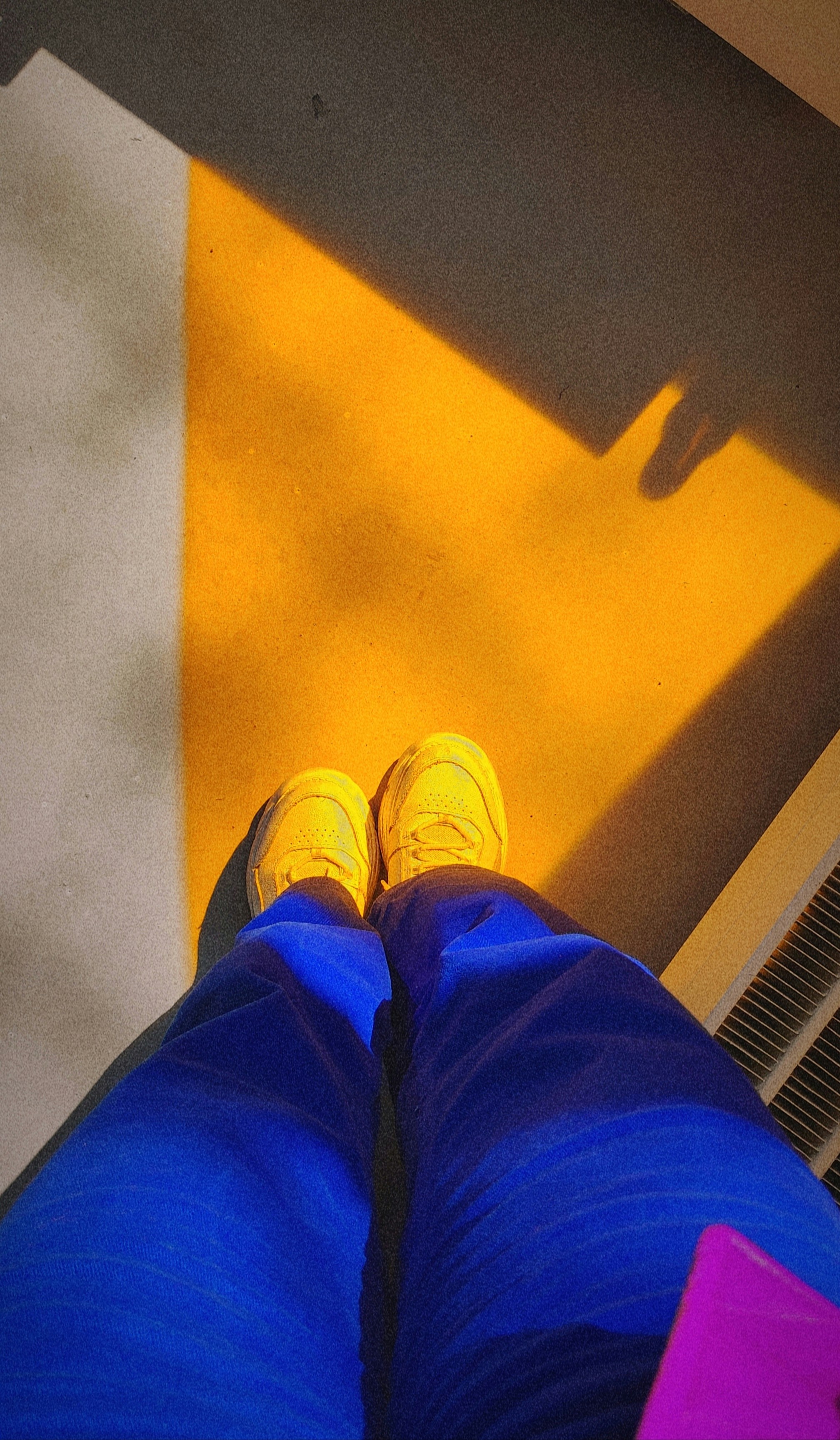 Top-down view of yellow sneakers worn over blue pants on a warm orange-tinted floor, with a vent visible to the right.