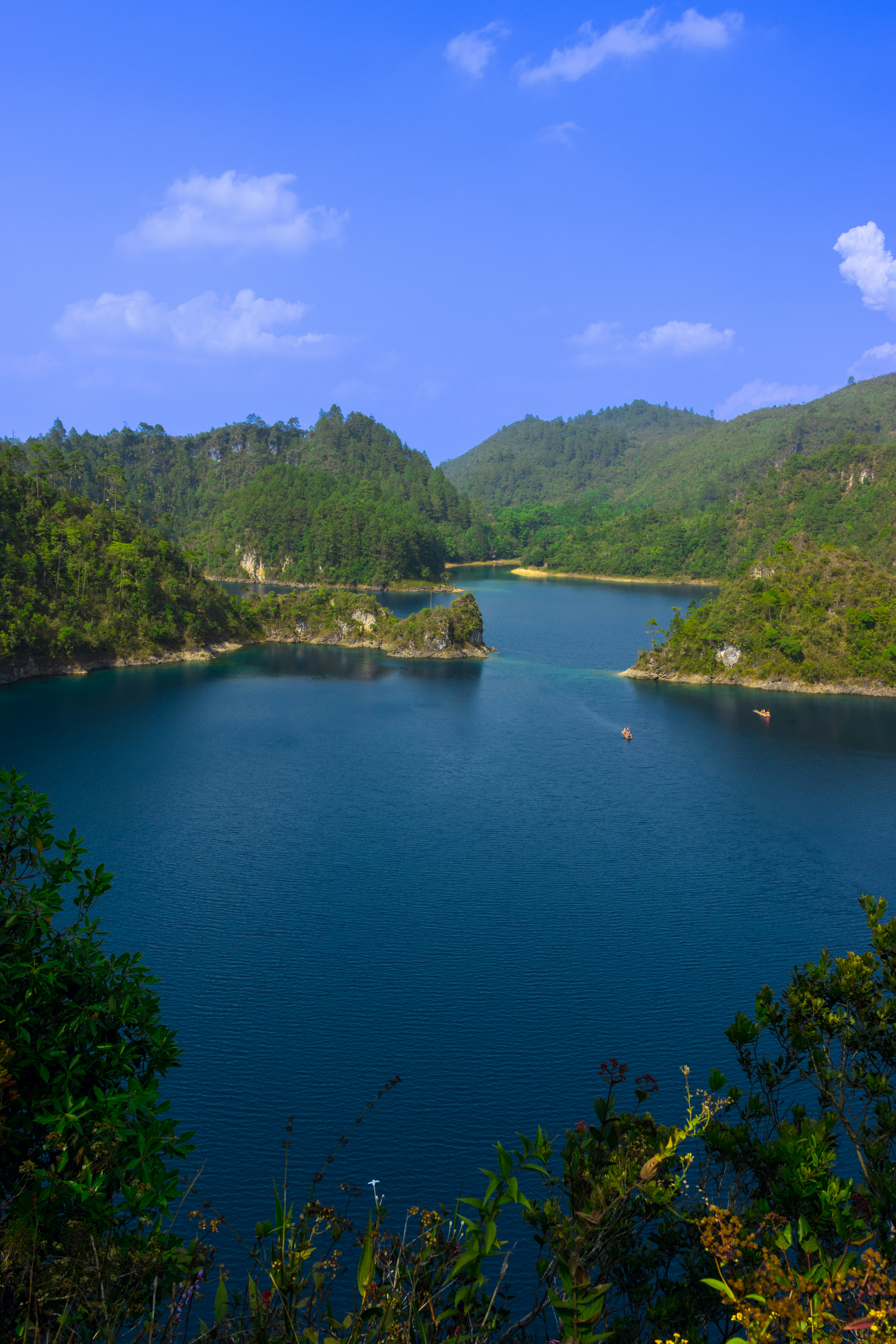 Mountainous landscape with a tranquil blue lake surrounded by lush greenery under a bright blue sky.