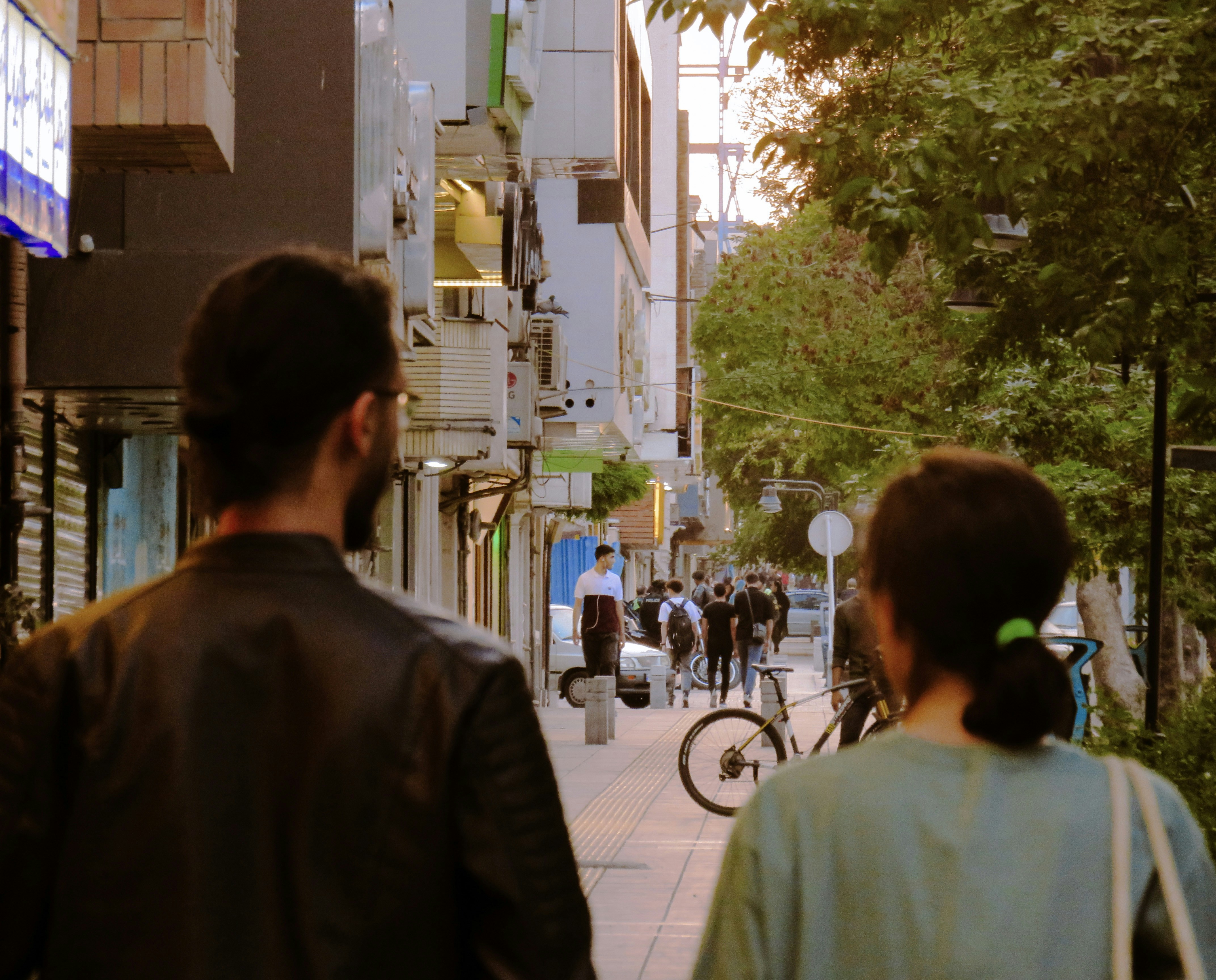 Two people walking along a tree-lined city street during sunset, with bicycles and buildings in the background.