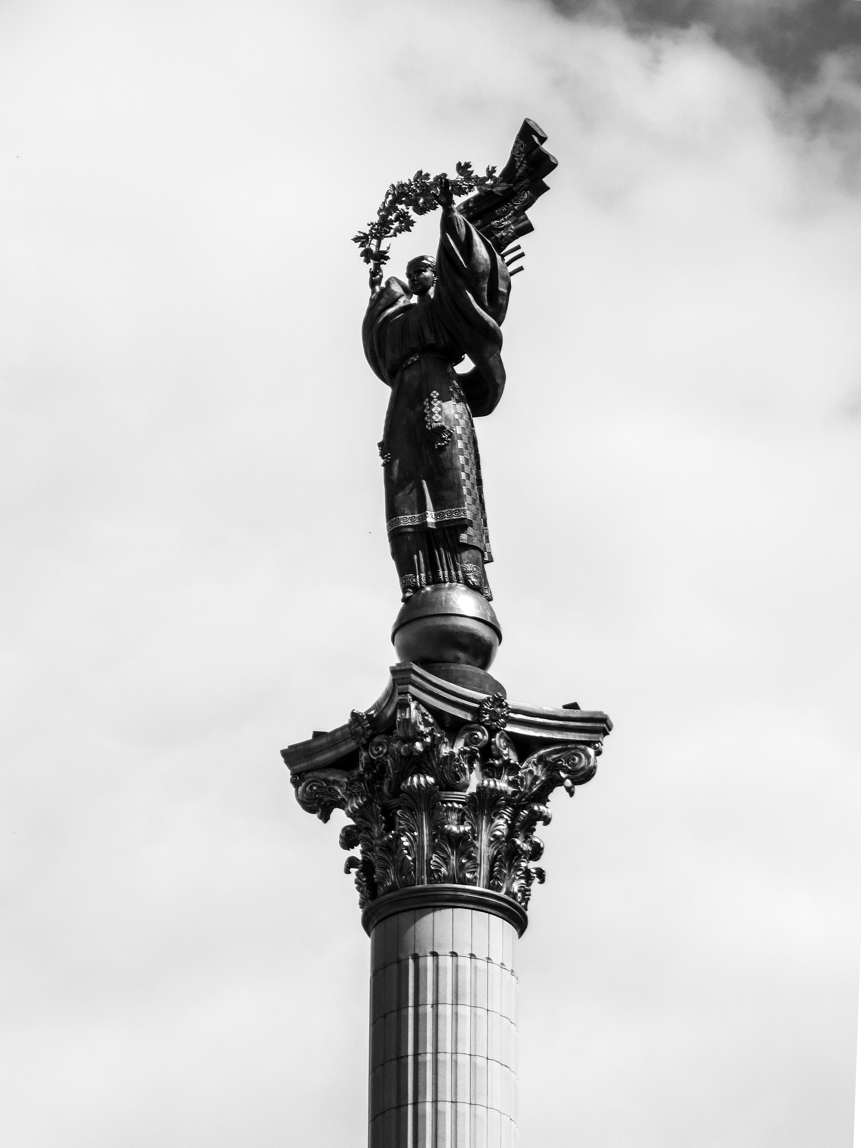 Monumental statue atop a tall column rises into a cloudy sky. Black-and-white photograph that emphasizes sculptural details and architectural grandeur.