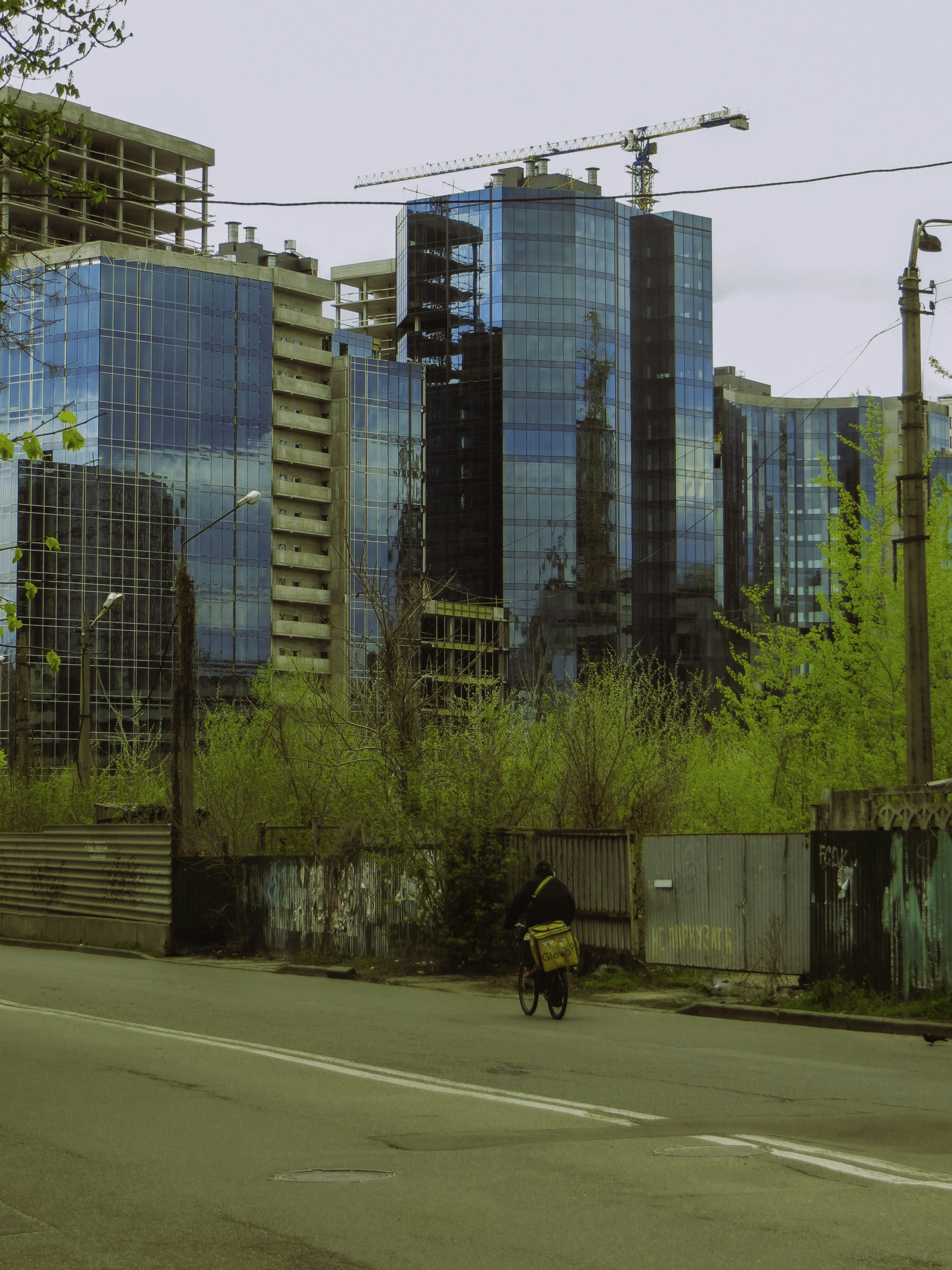 Glass-clad office towers under construction loom over a quiet street as a cyclist pedals by. A photograph capturing urban renewal in progress.