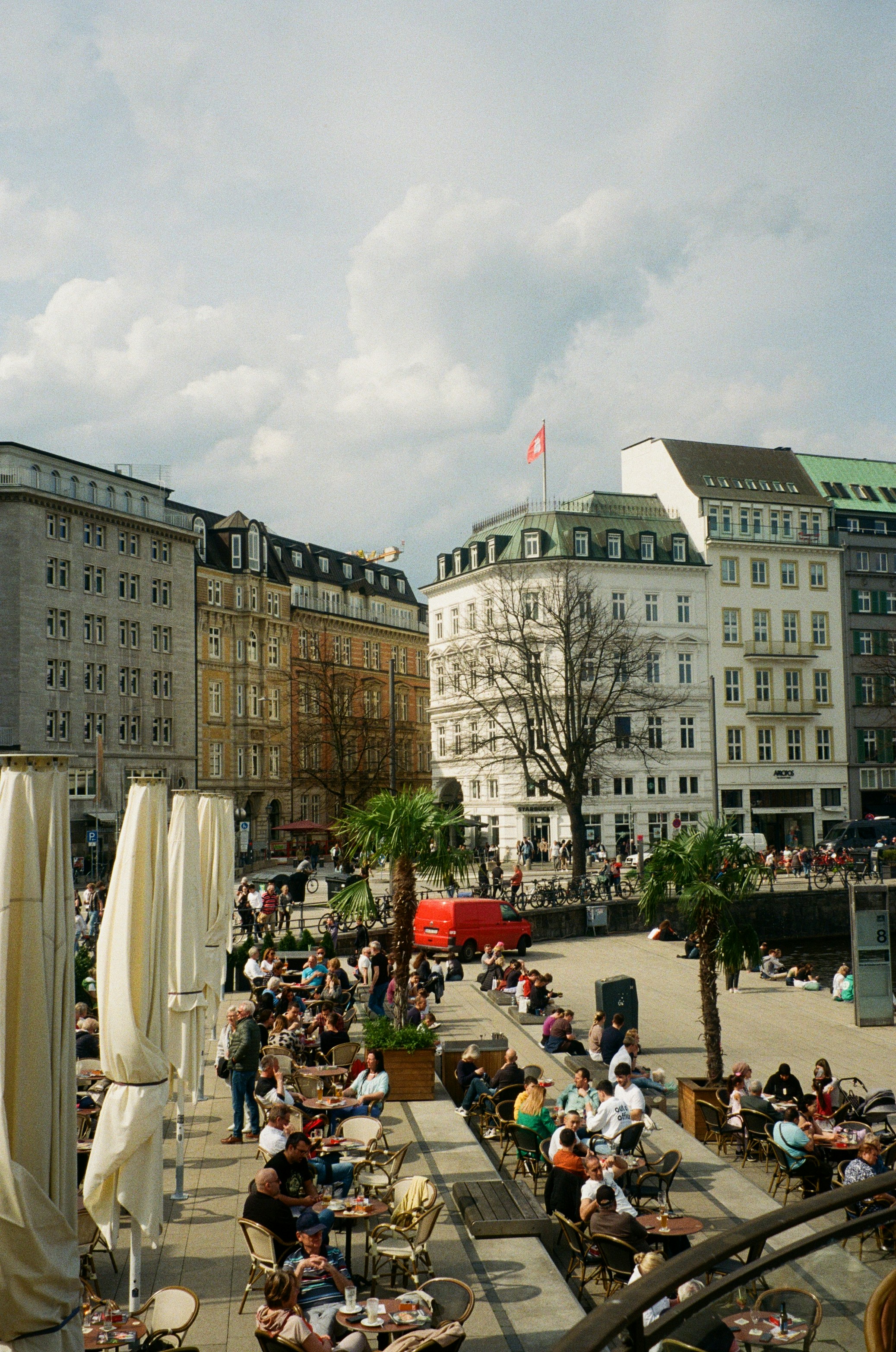 Outdoor cafe terrace with people dining along a pedestrian square, framed by colorful buildings and a bare tree under a bright sky.
