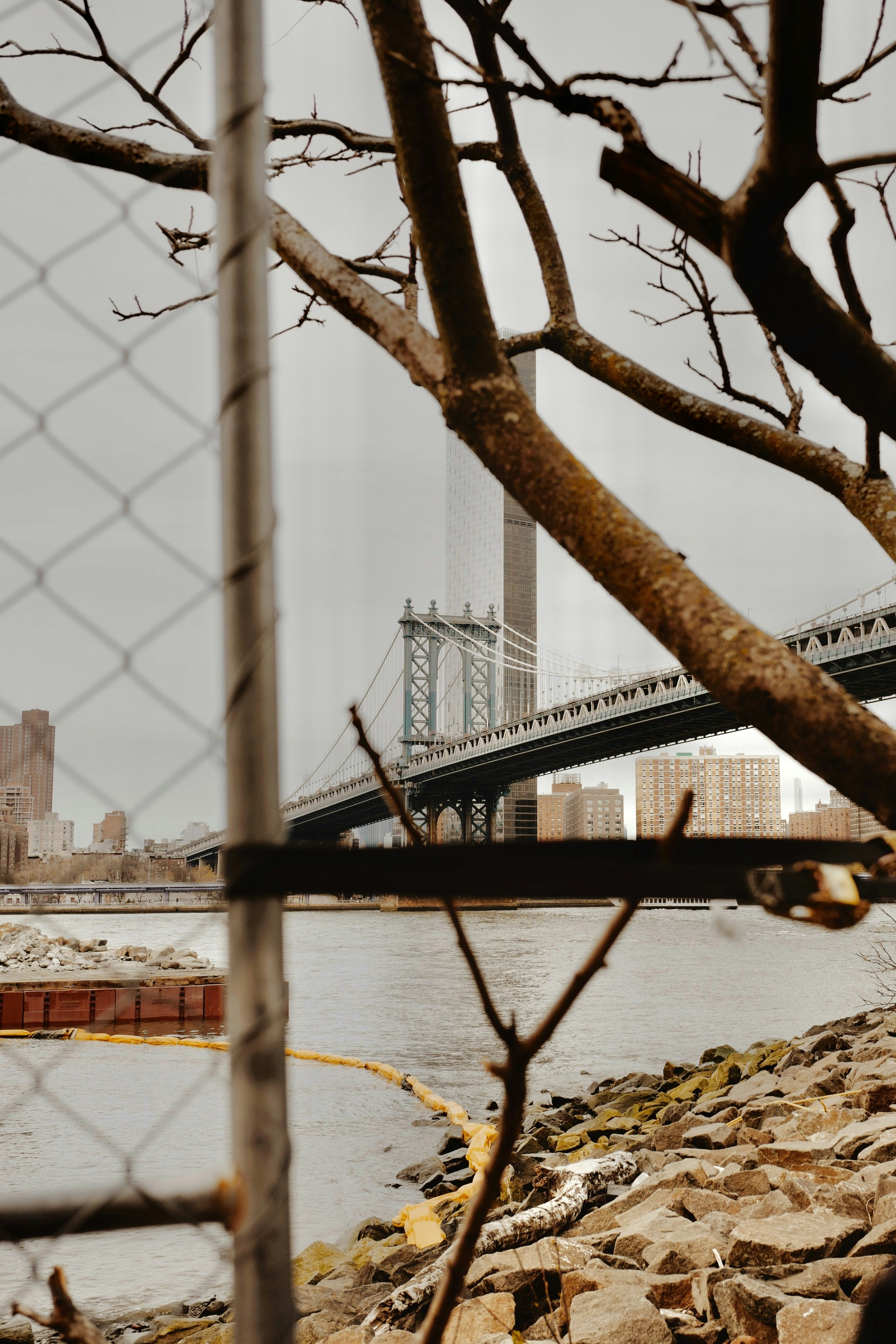 Chain-Link Veil: The Manhattan Bridge Framed by a Rocky Riverside
