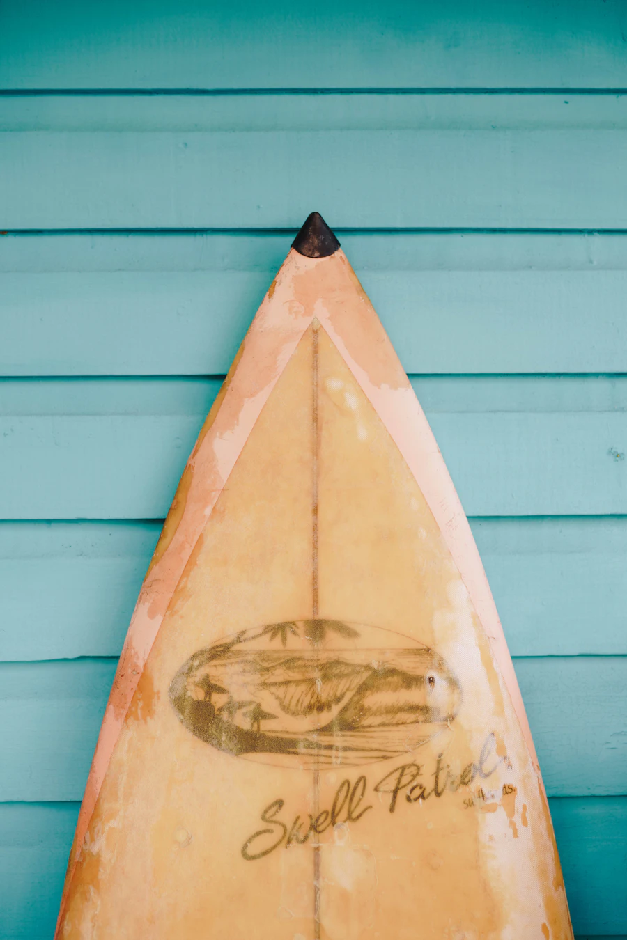 A weathered yellow surfboard with a faded logo leans against turquoise wooden planks, creating a beachy, vintage vibe.
