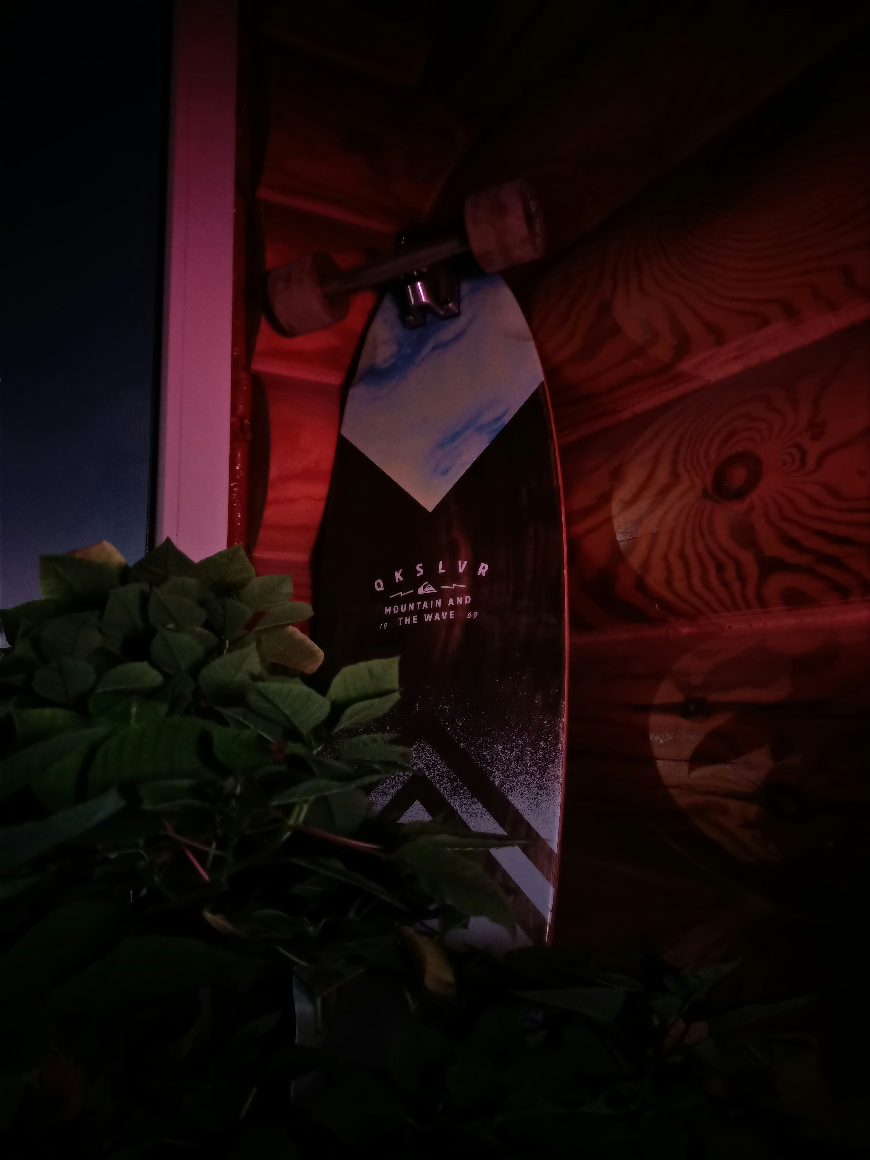 A snowboard leans against a wooden wall, bathed in moody red ambient light with green foliage in the foreground.