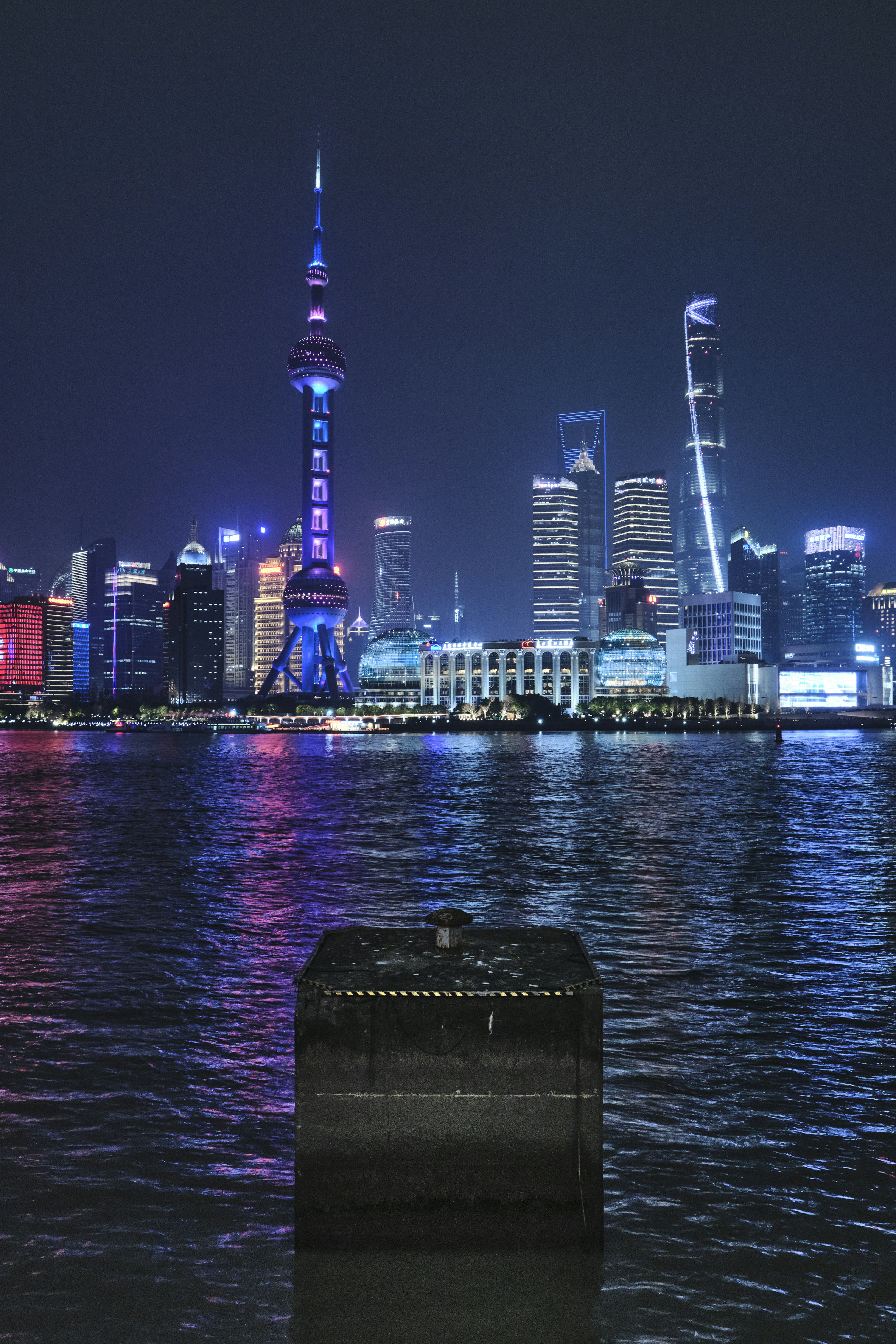 The iconic Oriental Pearl Tower stands illuminated against Shanghai's vibrant skyline, reflecting in the water below. A concrete pillar anchors the foreground.