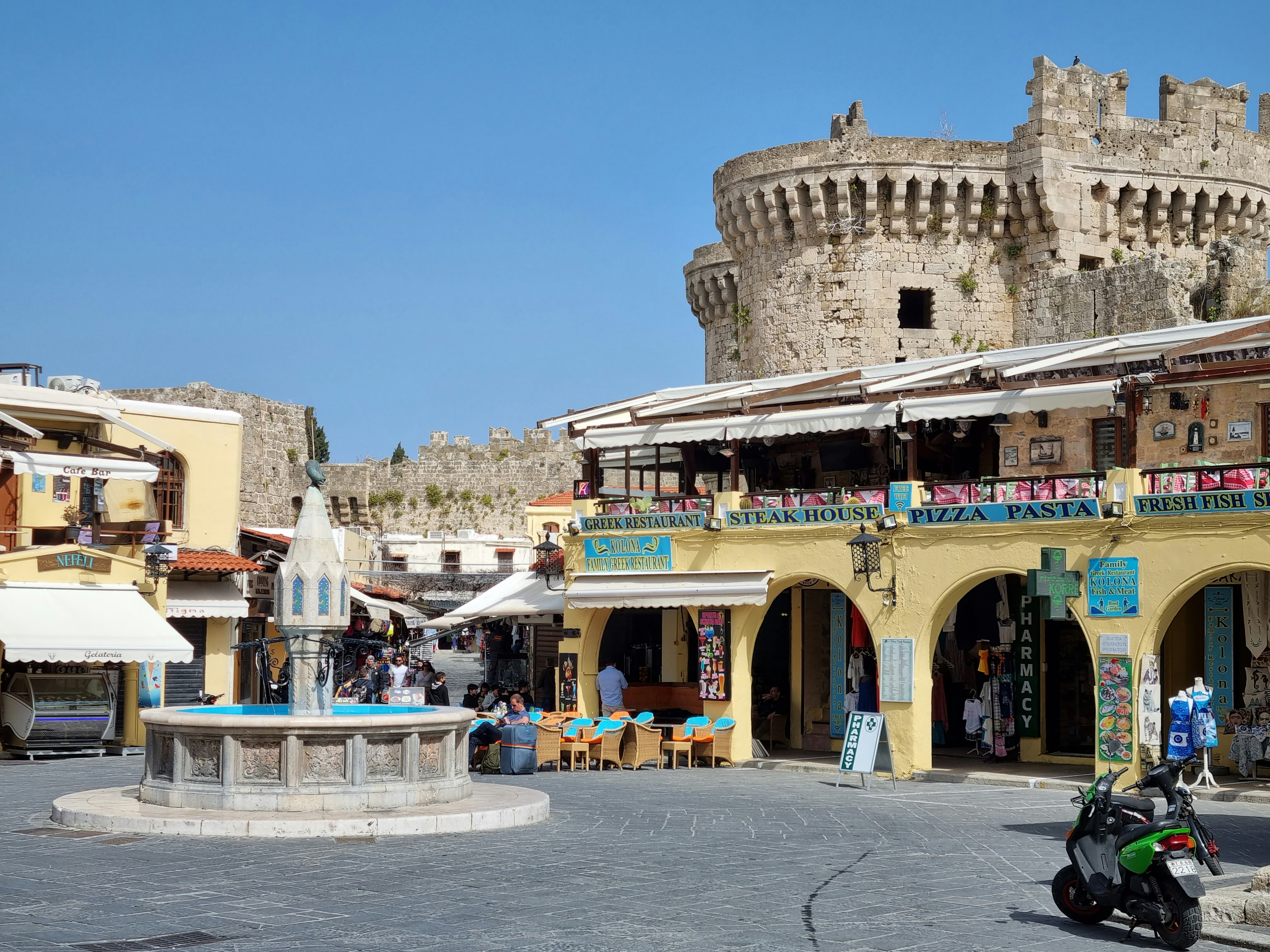 Bustling marketplace featuring colorful shops and a historic fortress backdrop. A central fountain adds charm to the lively scene.