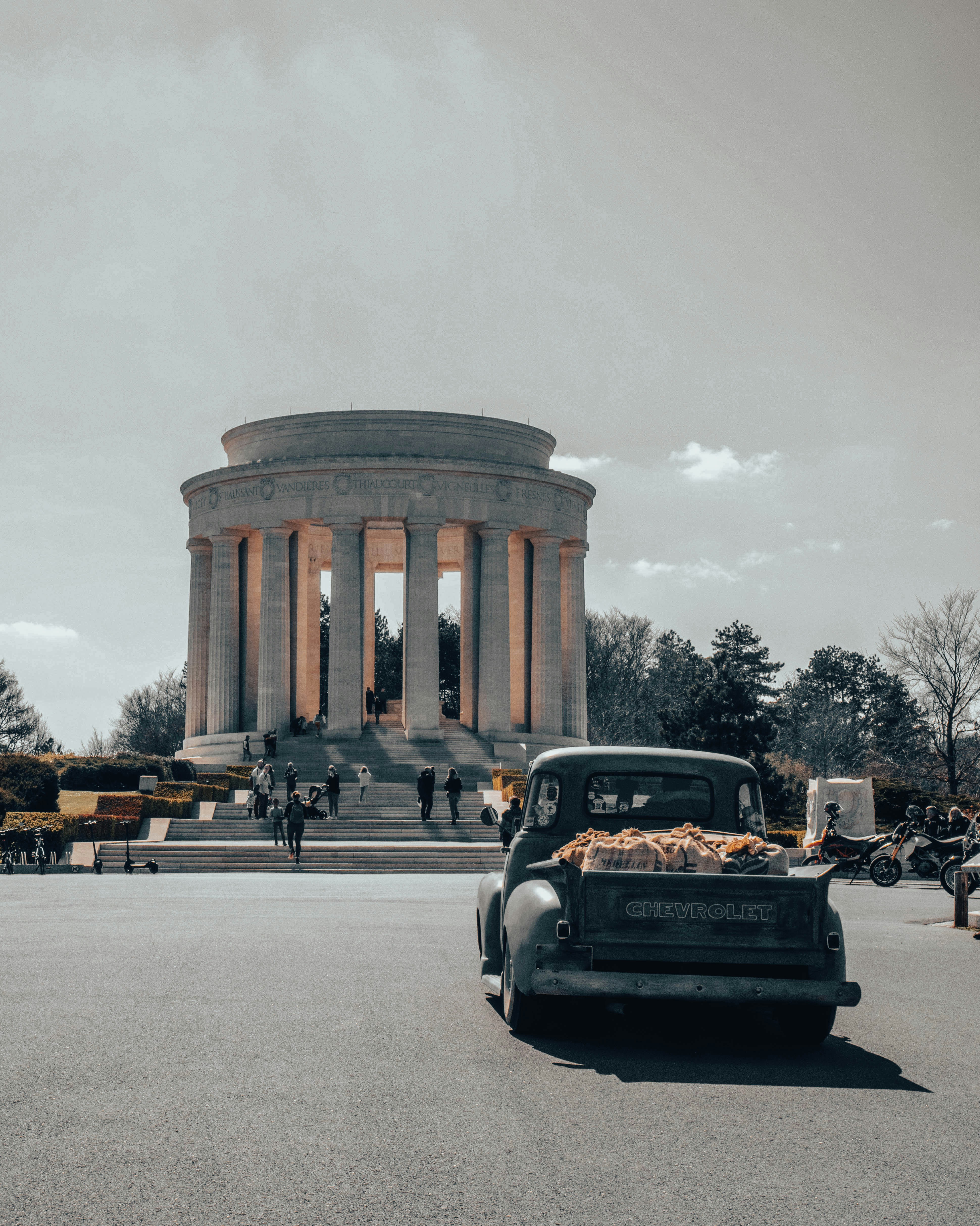 Vintage Chevy pickup rests on a wide road in front of a neoclassical temple, with visitors on the steps and surrounding park. This color photograph captures a quiet moment where eras collide.