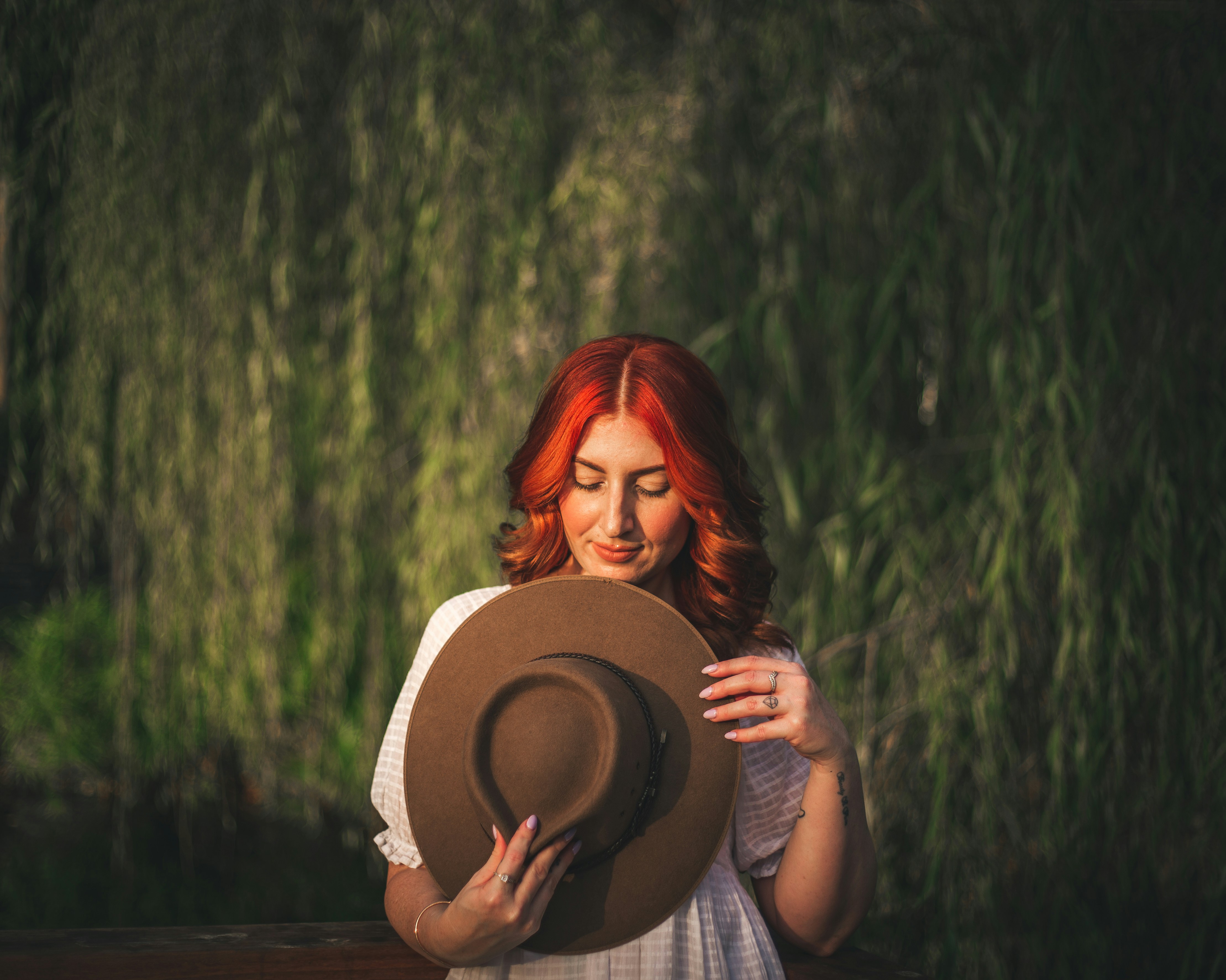 Woman with red hair holding a brown hat, set against lush green foliage.