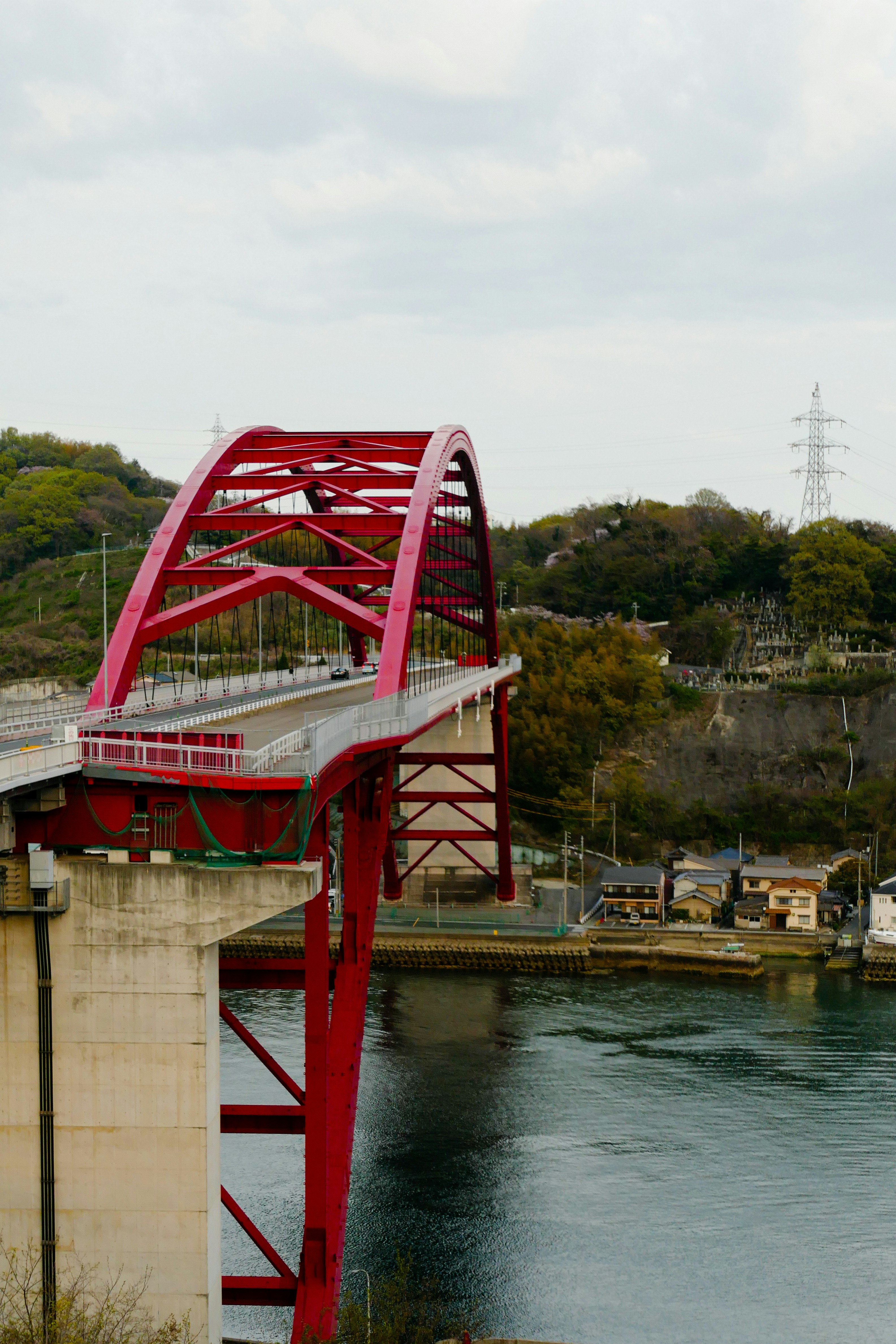 Red arch bridge spanning a wide river with surrounding greenery and overcast sky.
