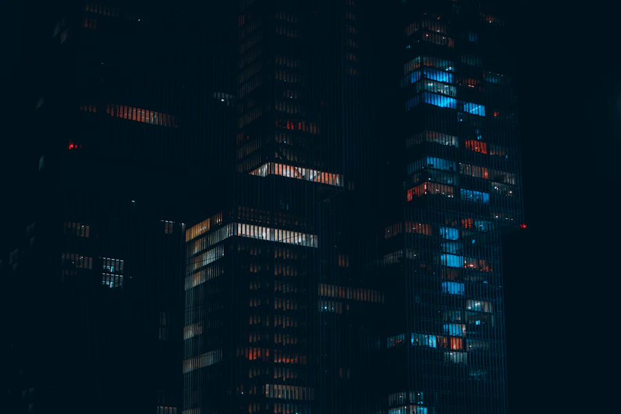 Dark city skyscrapers with scattered lit windows and blue-red office lights glowing against the night sky.