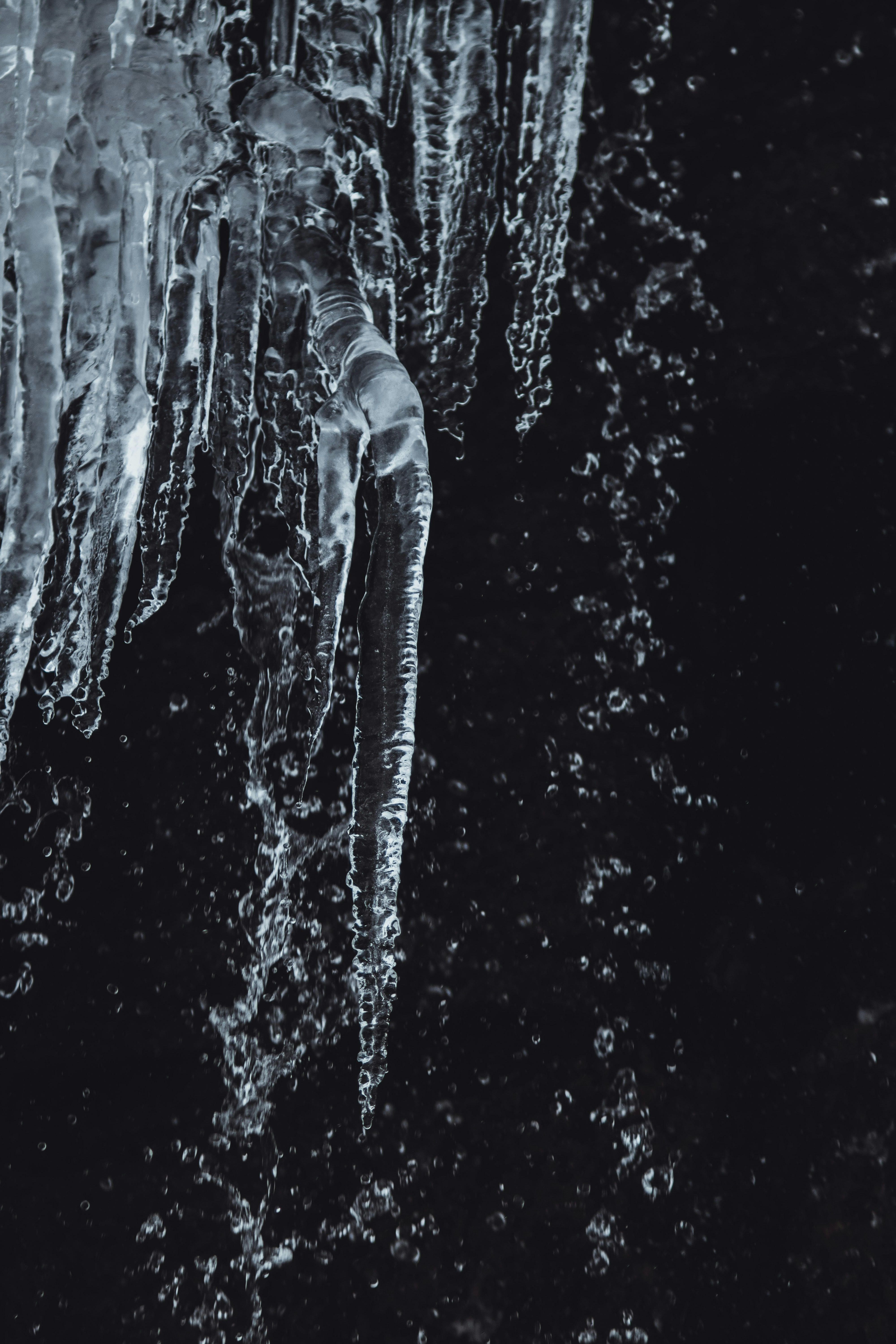 Icicles hanging from a dark surface, showcasing intricate details and textures. Water droplets cascade around them, enhancing the scene's dynamic contrast.