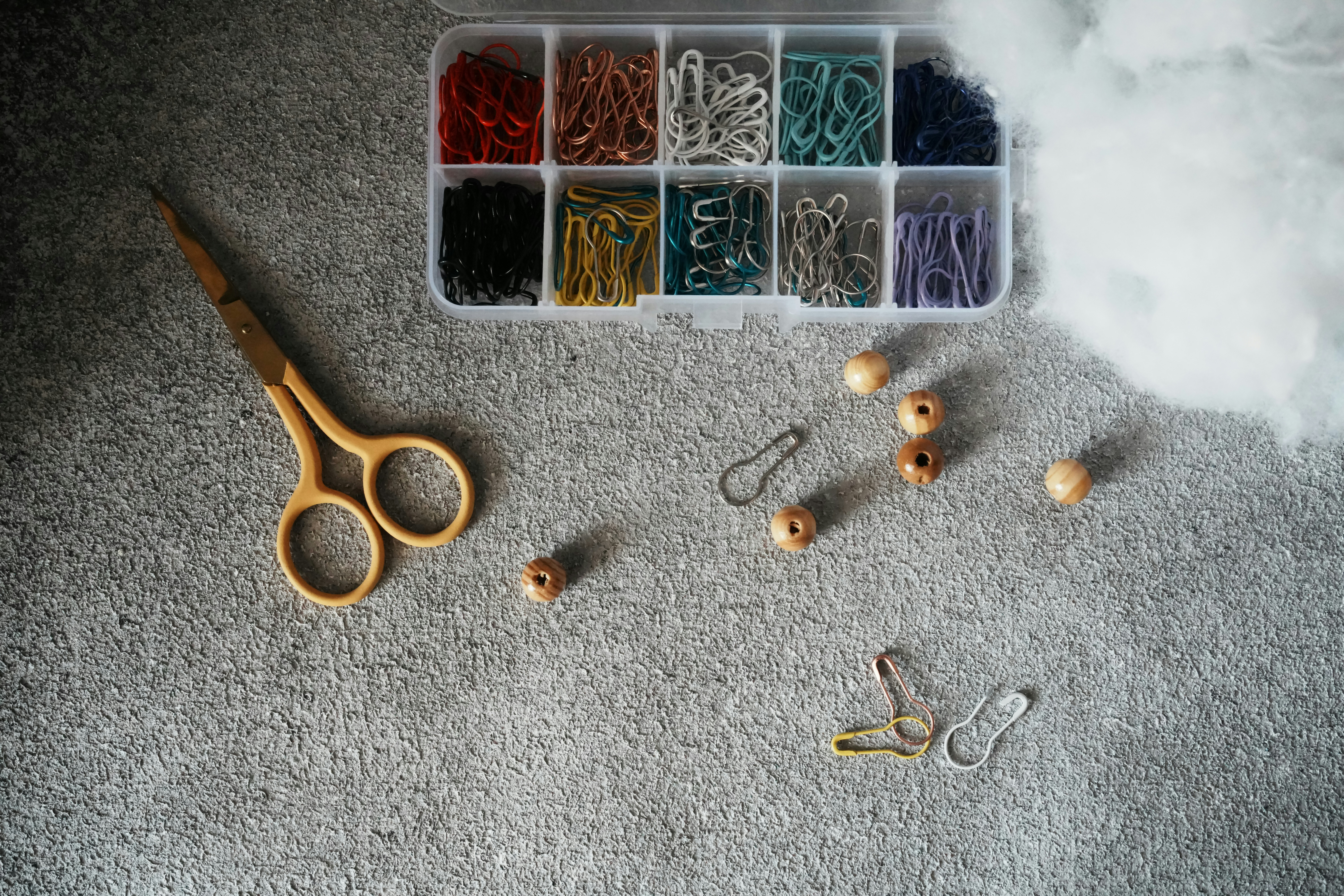 A collection of crafting materials including scissors, colorful paper clips, wooden beads, and fluffy cotton on a textured surface.
