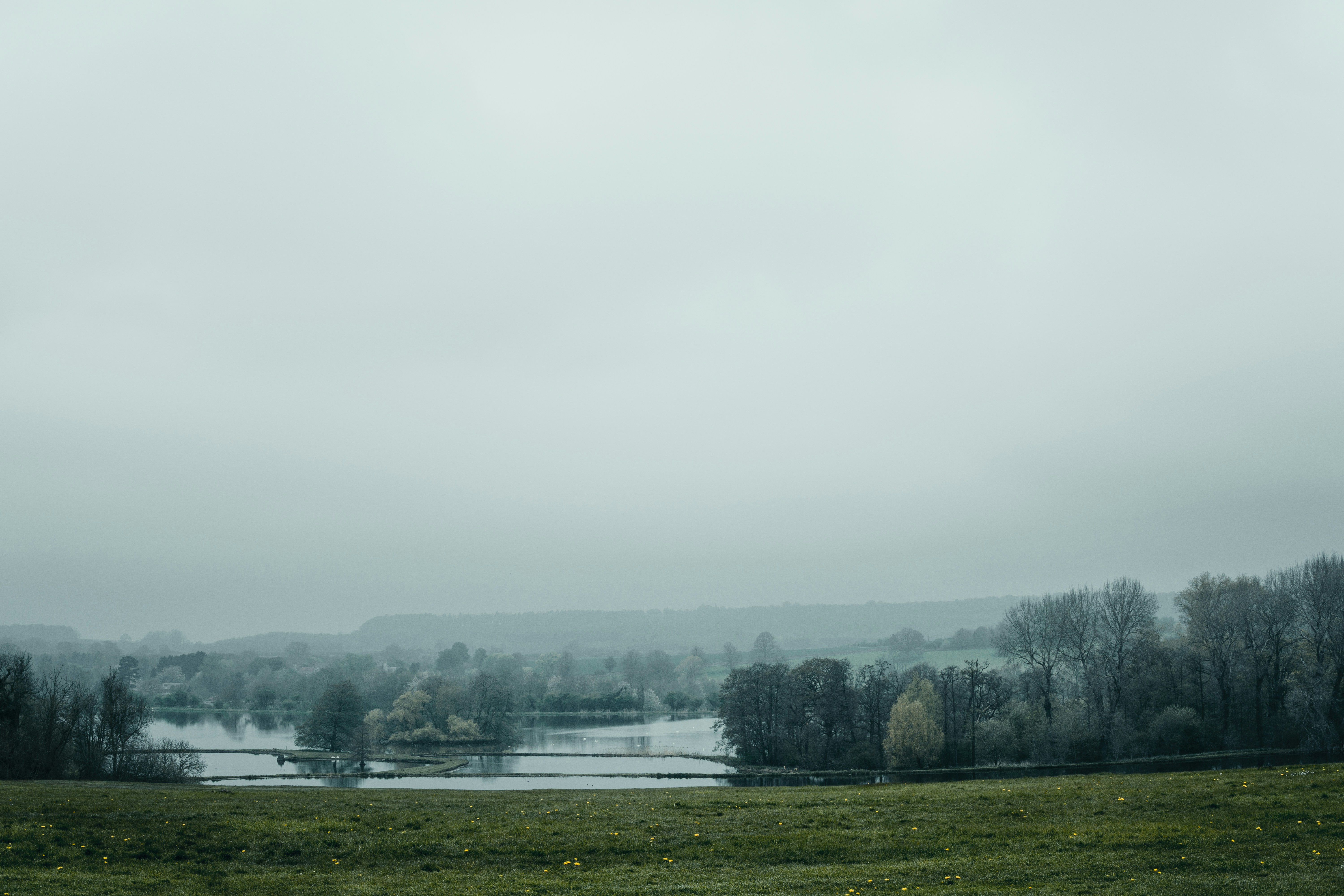 Overcast sky above a tranquil lake surrounded by lush green fields and distant trees.