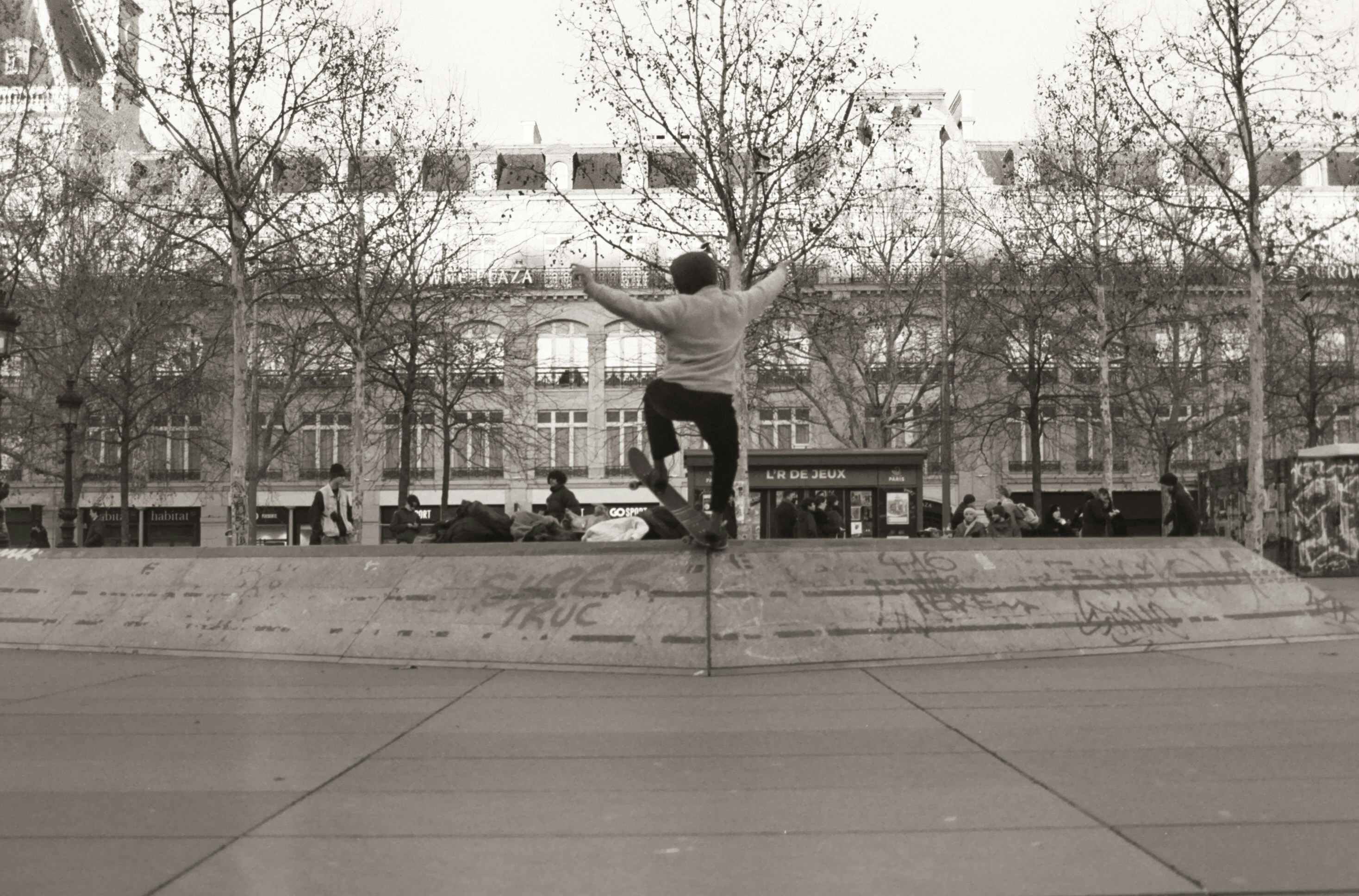 Black-and-white photograph of a skater grinding a curved rail in a city square. Leafless trees and urban facades frame the moment, emphasizing motion and balance.