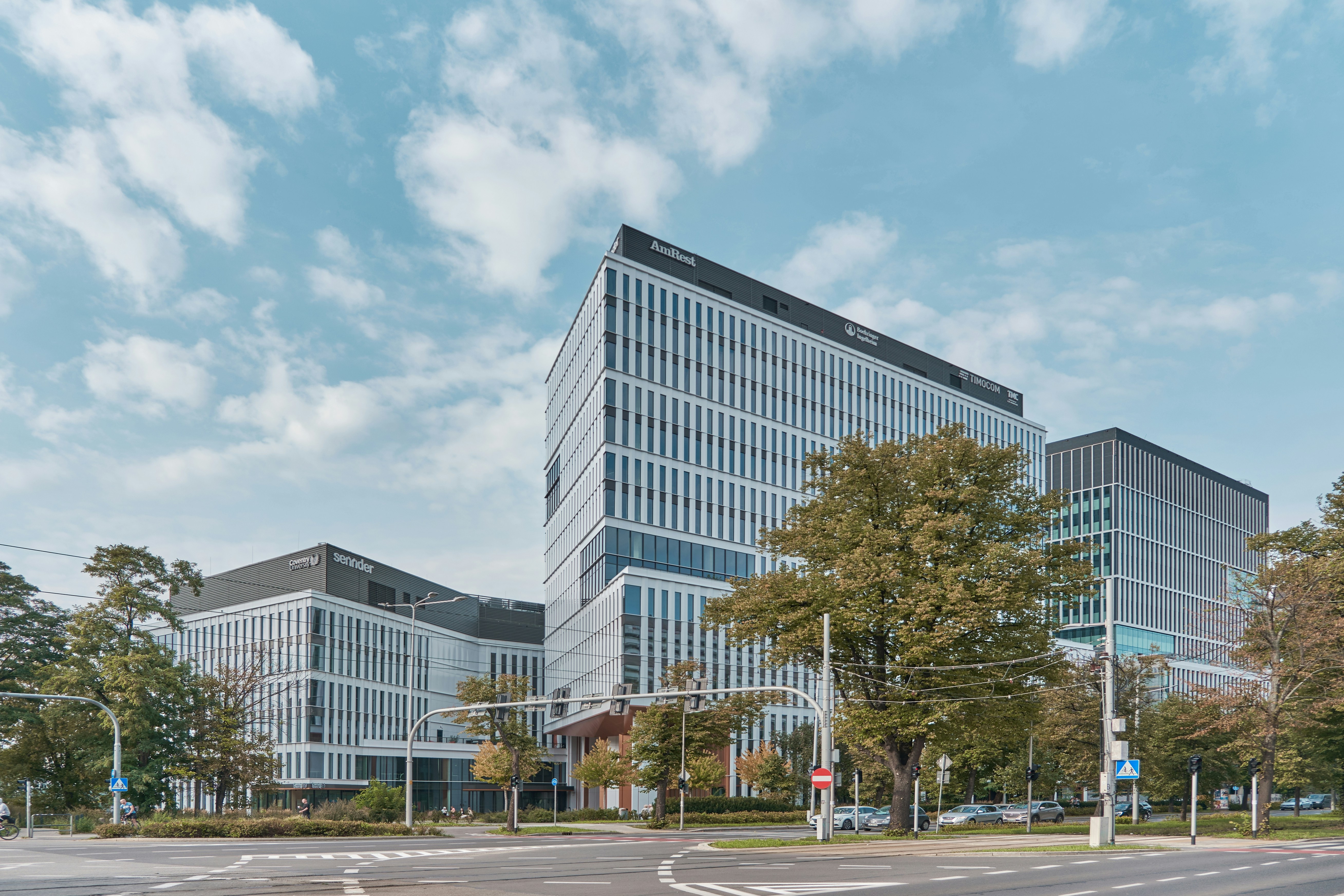 Contemporary office building with glass façade at a street intersection, surrounded by trees and under a partly cloudy sky.