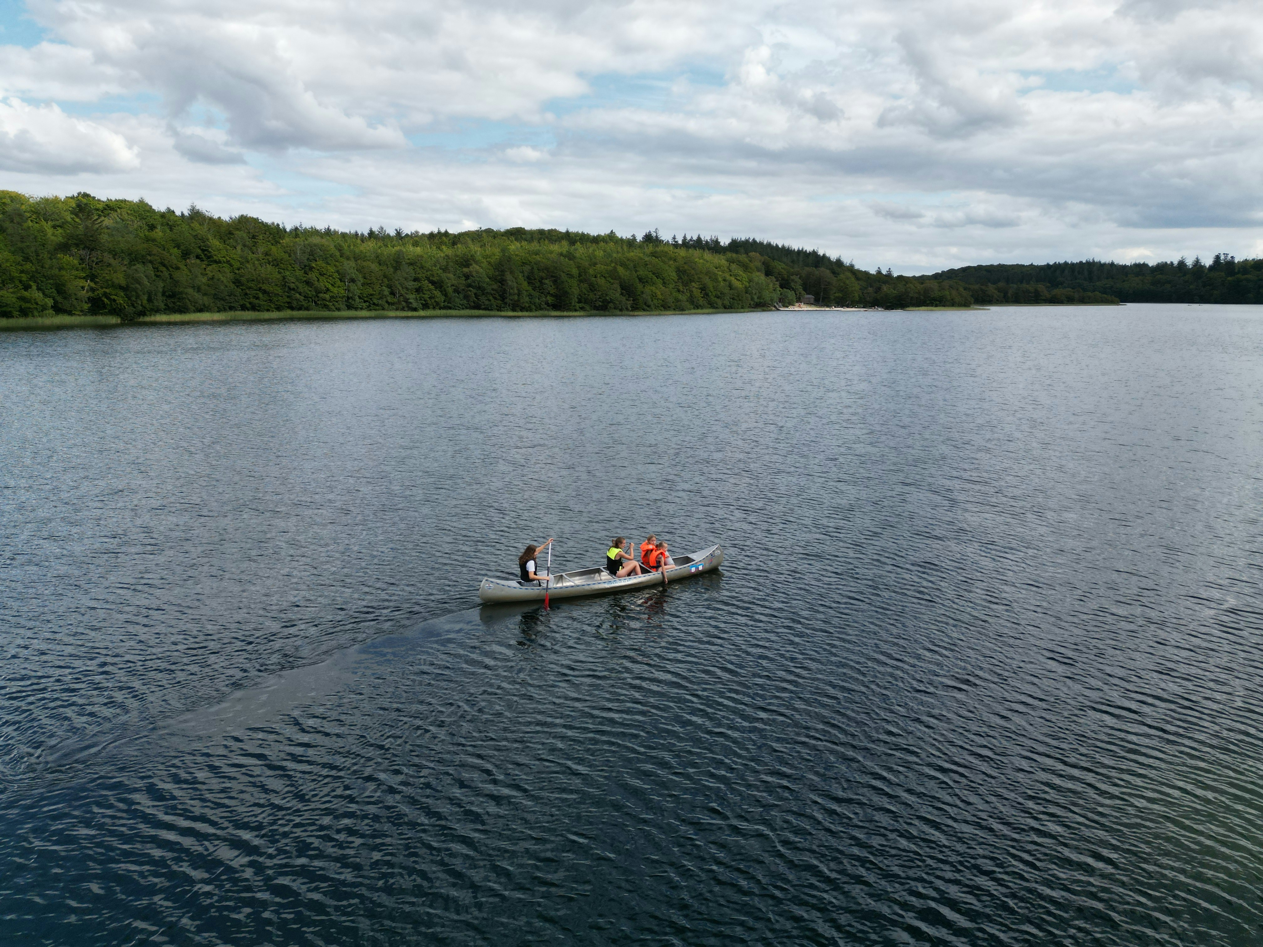 Three individuals paddling a canoe on a calm lake surrounded by lush greenery and a cloudy sky.