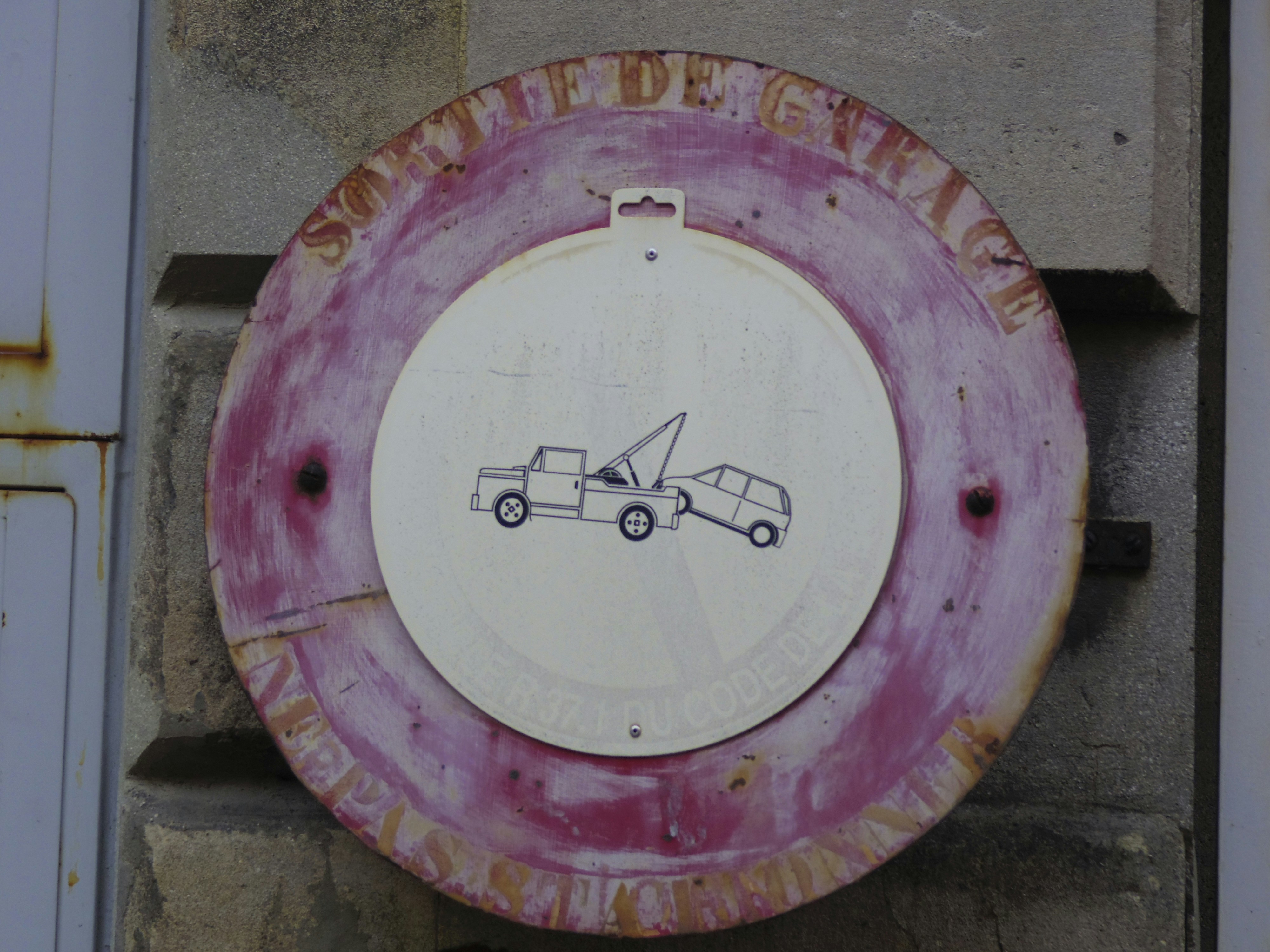 Weathered circular sign mounted on a brick wall features an illustration of a tow truck towing a car, indicating a tow-away zone. The purple ring and worn surface emphasize an urban signage element.