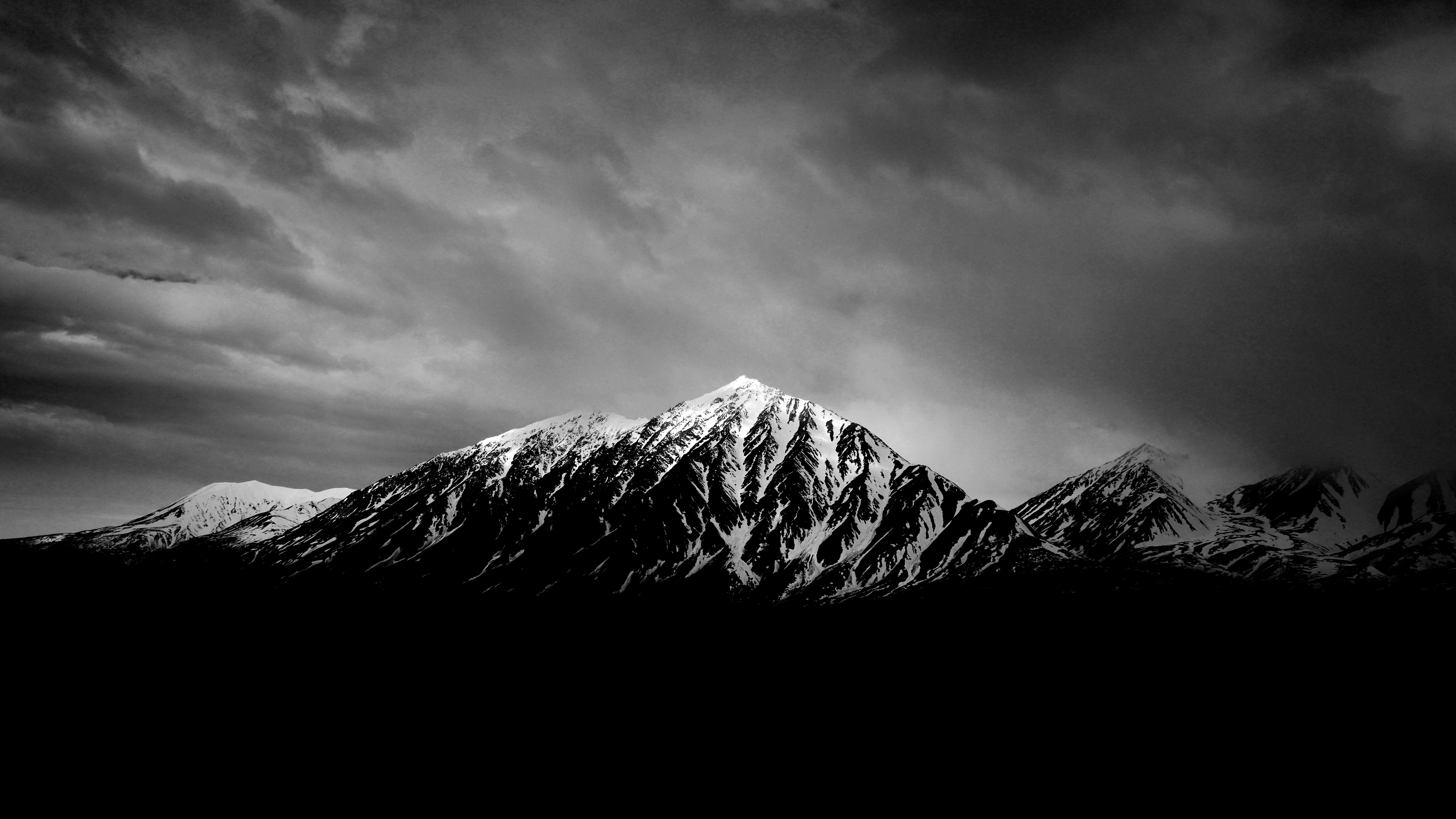 Snow-capped mountain range under dramatic cloudy sky in black and white.