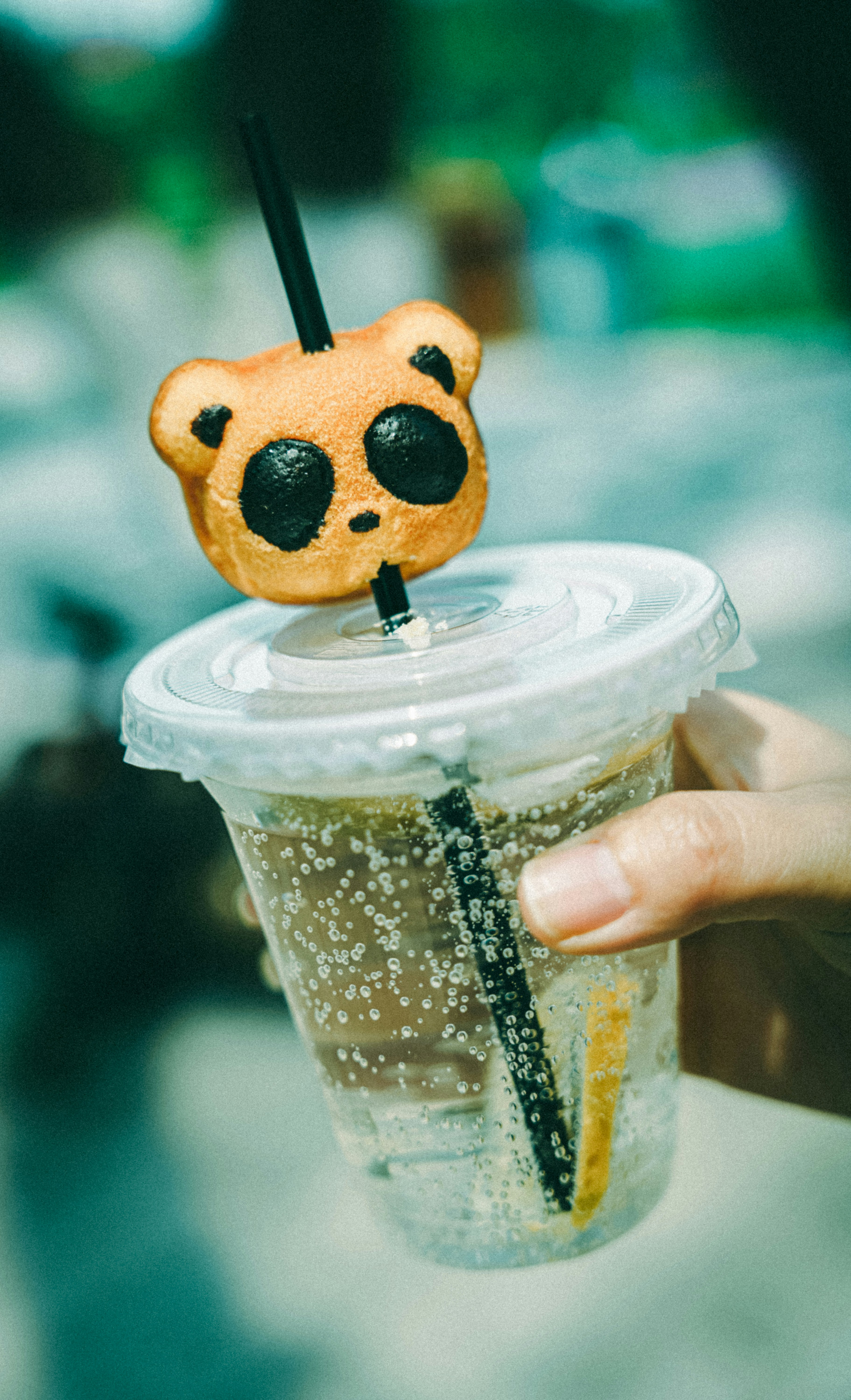 A hand holding a clear cup of sparkling beverage topped with a panda-shaped cookie. The scene conveys a lighthearted and fun atmosphere.