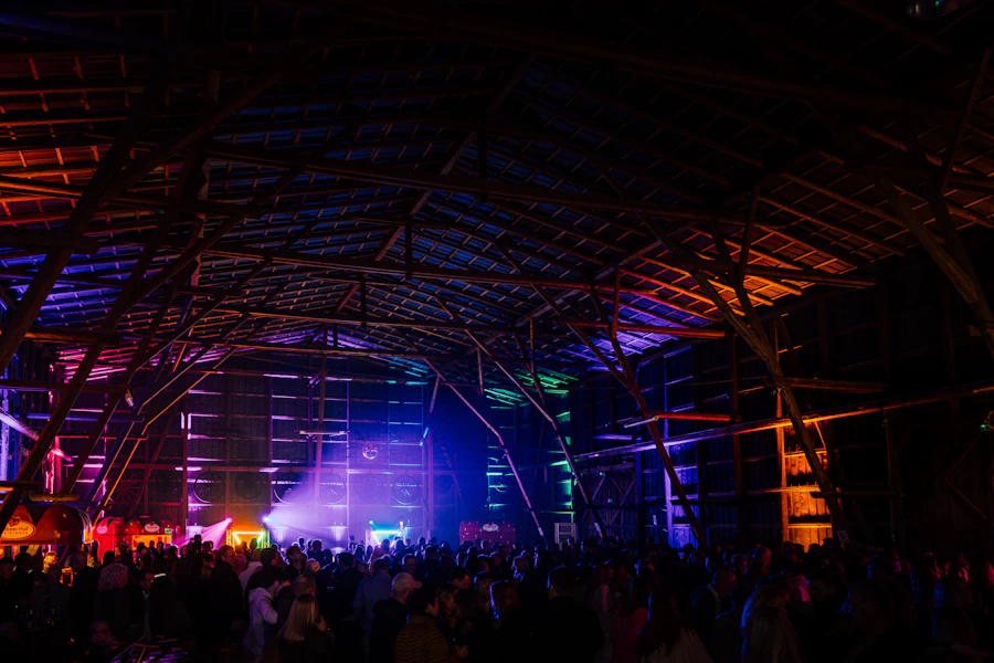 A packed crowd gathers inside a dark warehouse venue lit by vivid purple, blue, and orange stage lights. Metal roof trusses and glowing beams frame the dance floor.