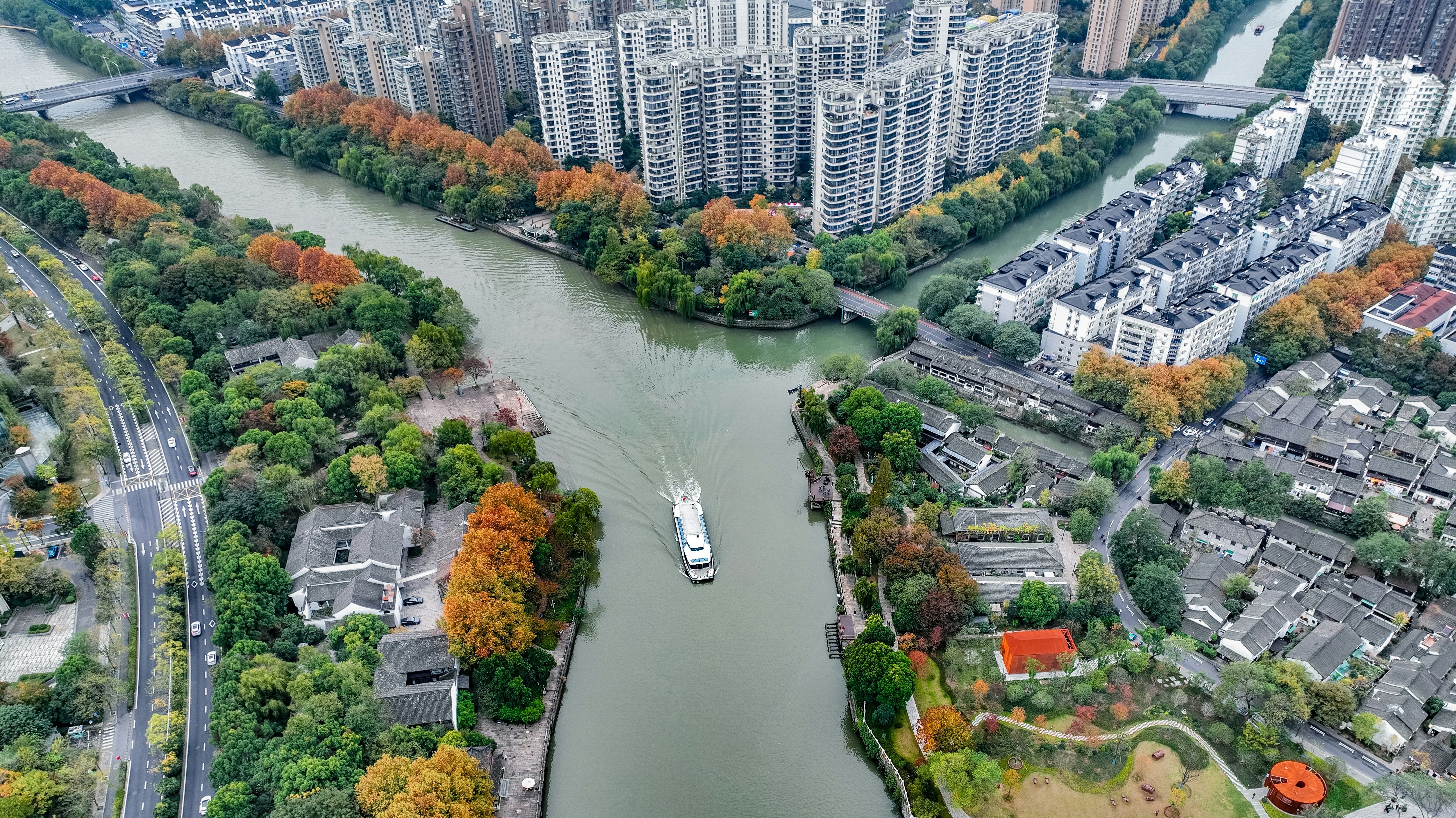 Aerial view of a river merging with urban surroundings, showcasing autumn foliage and high-rise buildings.
