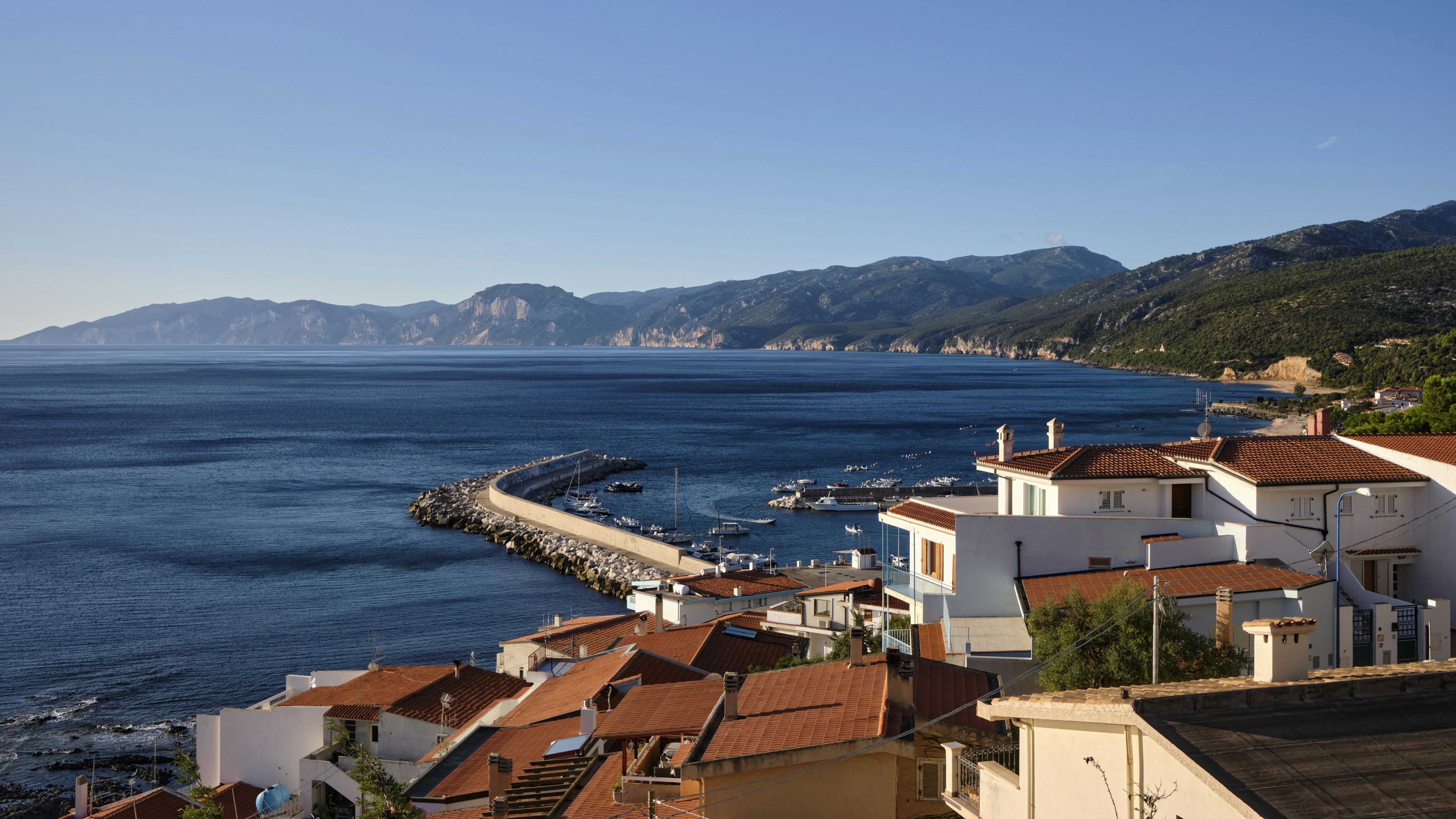 Terracotta rooftops overlook a tranquil harbor with a distant mountain range under a clear blue sky.