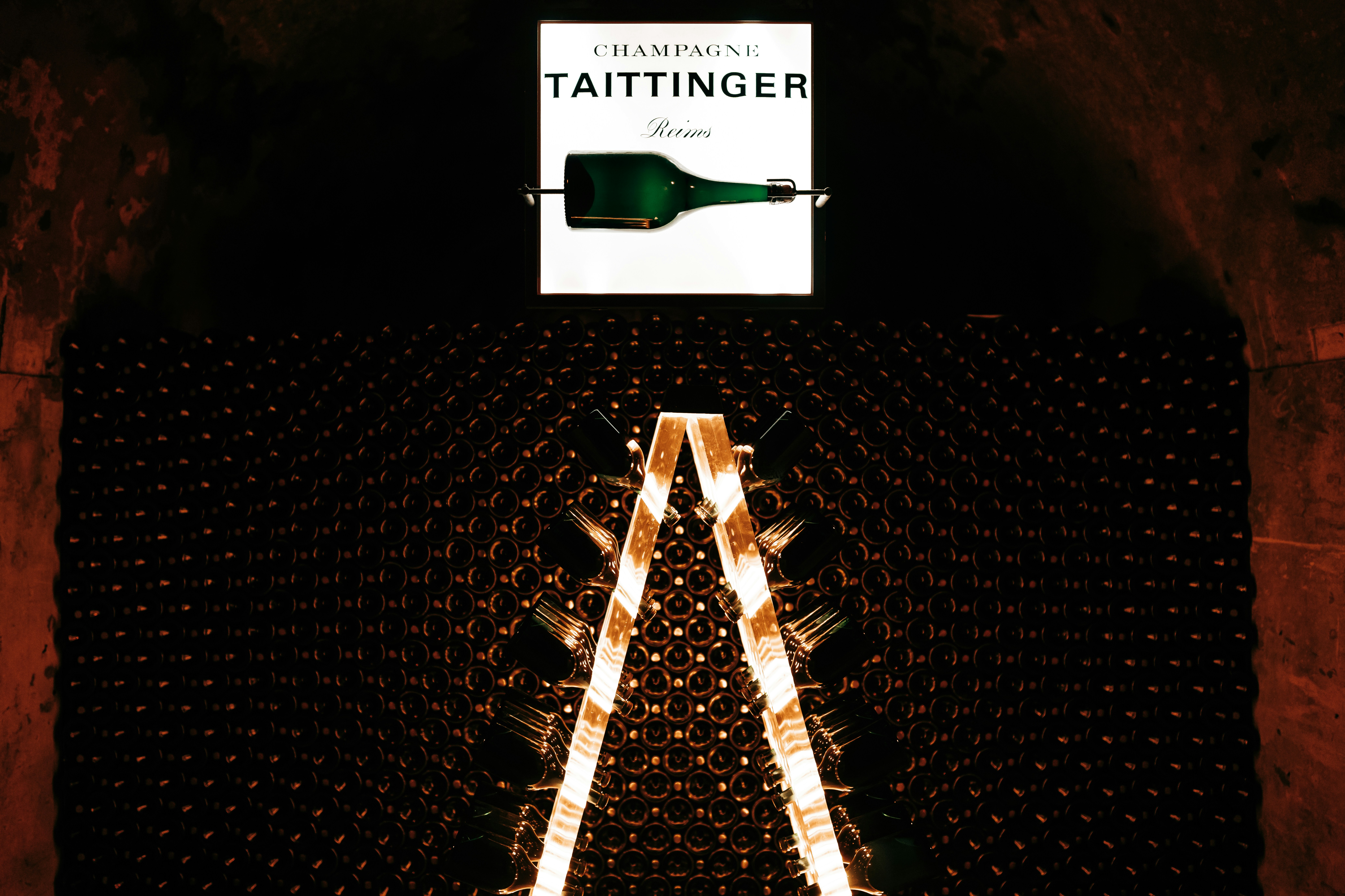 A striking arrangement of champagne bottles forming an illuminated triangular pattern beneath a Taittinger sign.