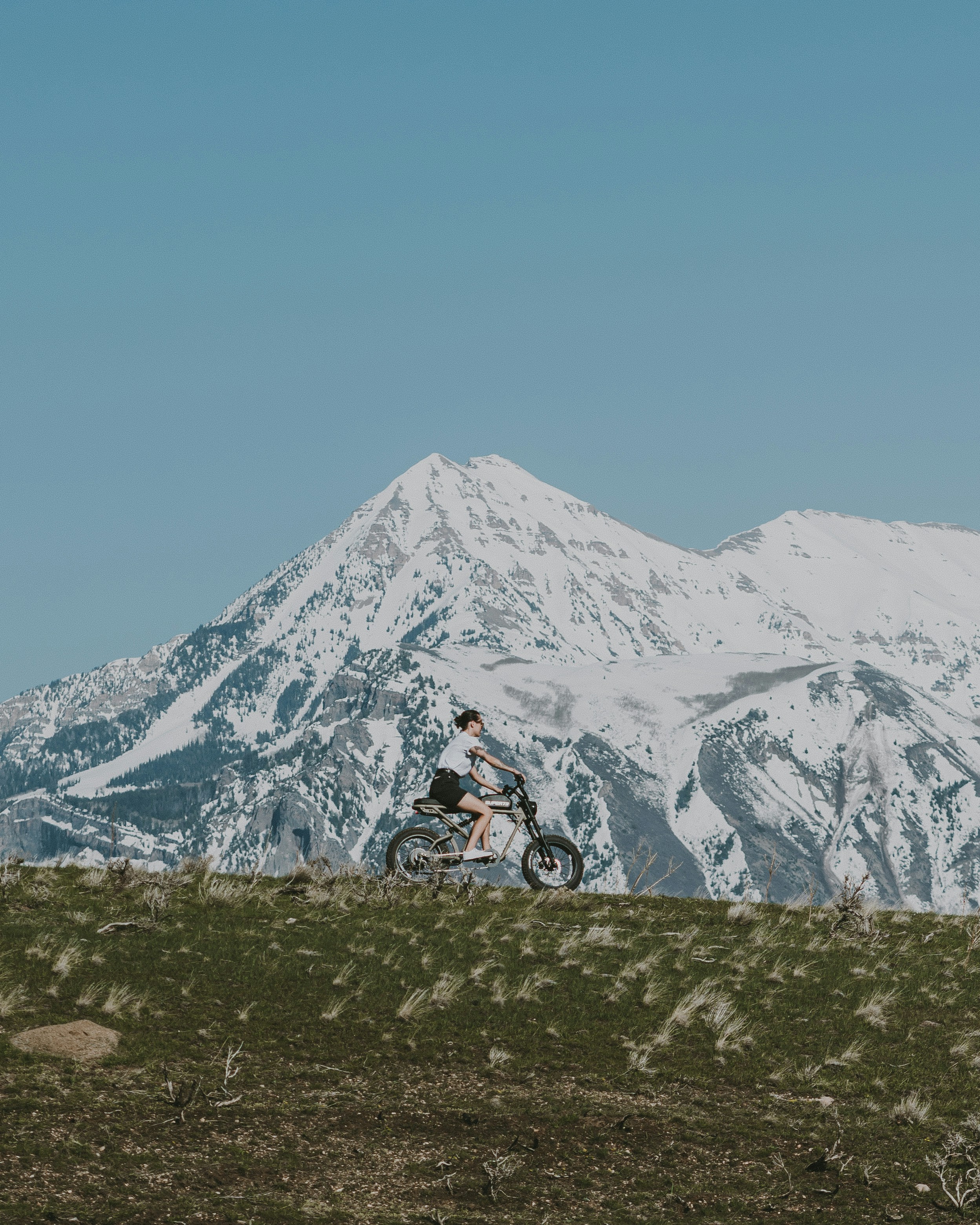 Ridgebound Rider: A lone cyclist crossing a grassy crest before snowbound giants