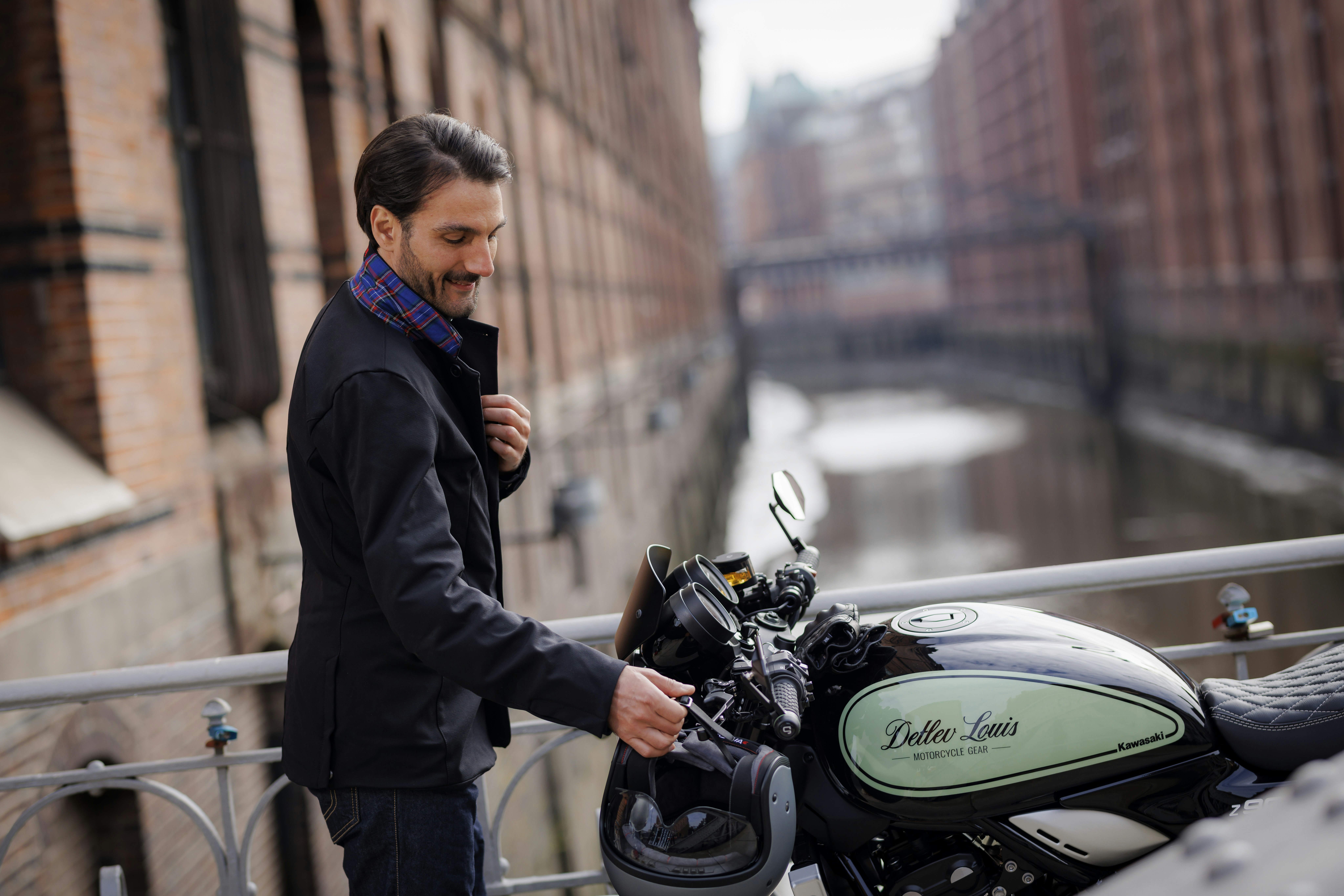 Man in a jacket standing beside a motorcycle on a bridge over a canal with historic brick buildings in the background.