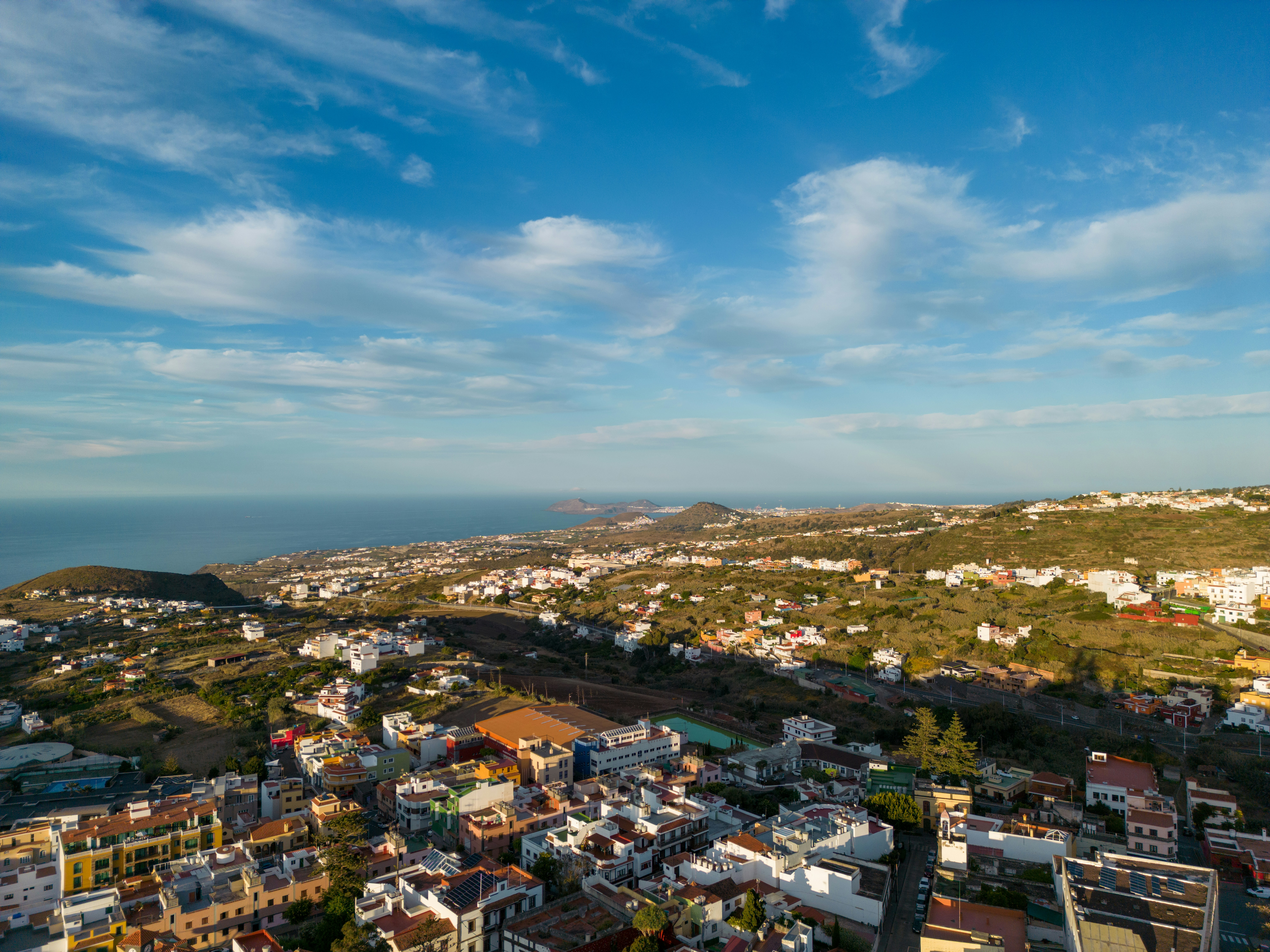 Aerial view of a coastal town with sprawling buildings and green landscapes under a partly cloudy sky.