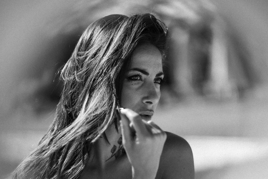 Black-and-white portrait of a woman in profile with windblown hair, looking off to the right. Soft blurred background keeps attention on her face and hand.