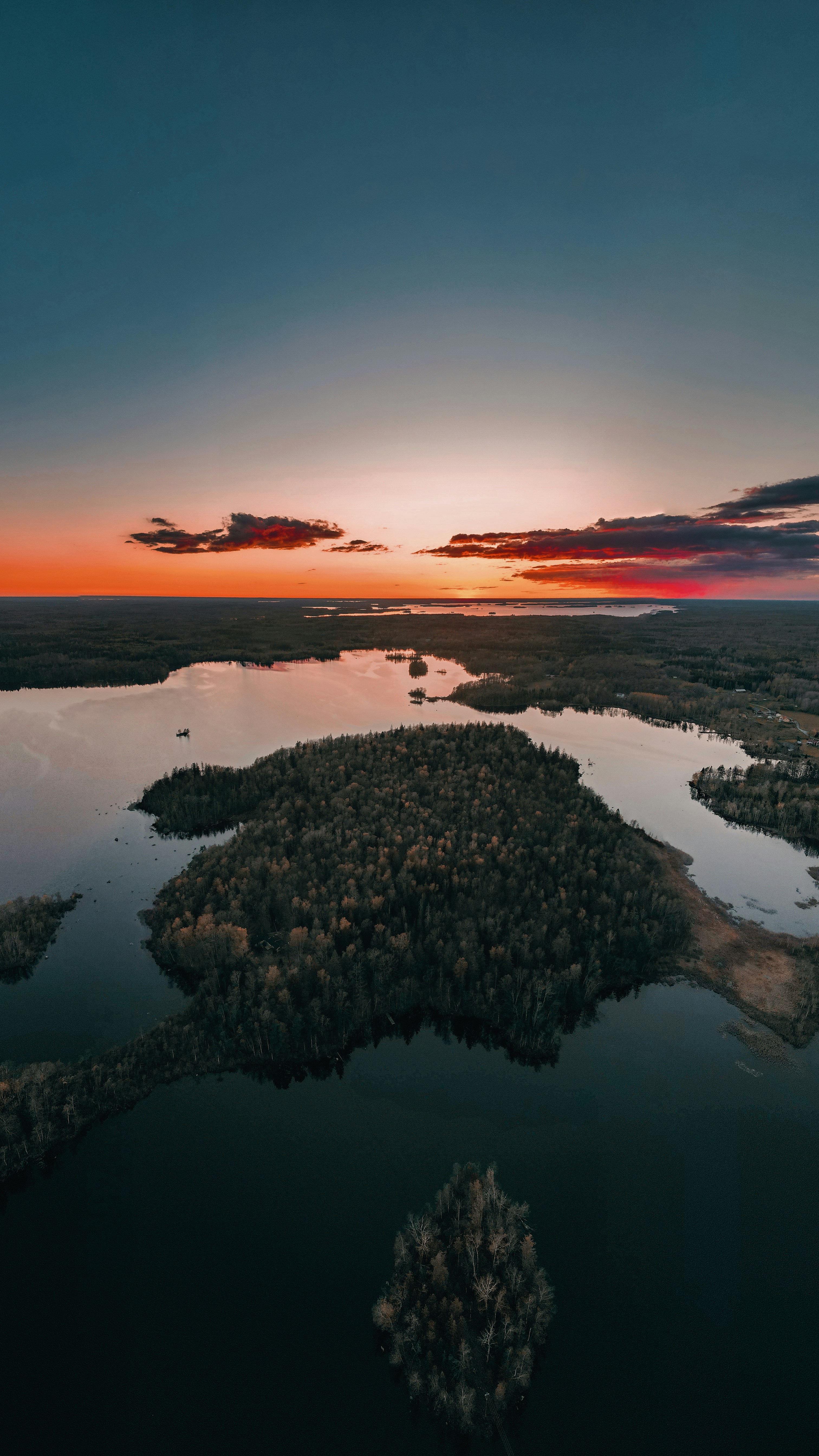 Aerial view of a tranquil landscape featuring an island surrounded by reflective waters at twilight. The horizon glows with soft hues of orange and purple.