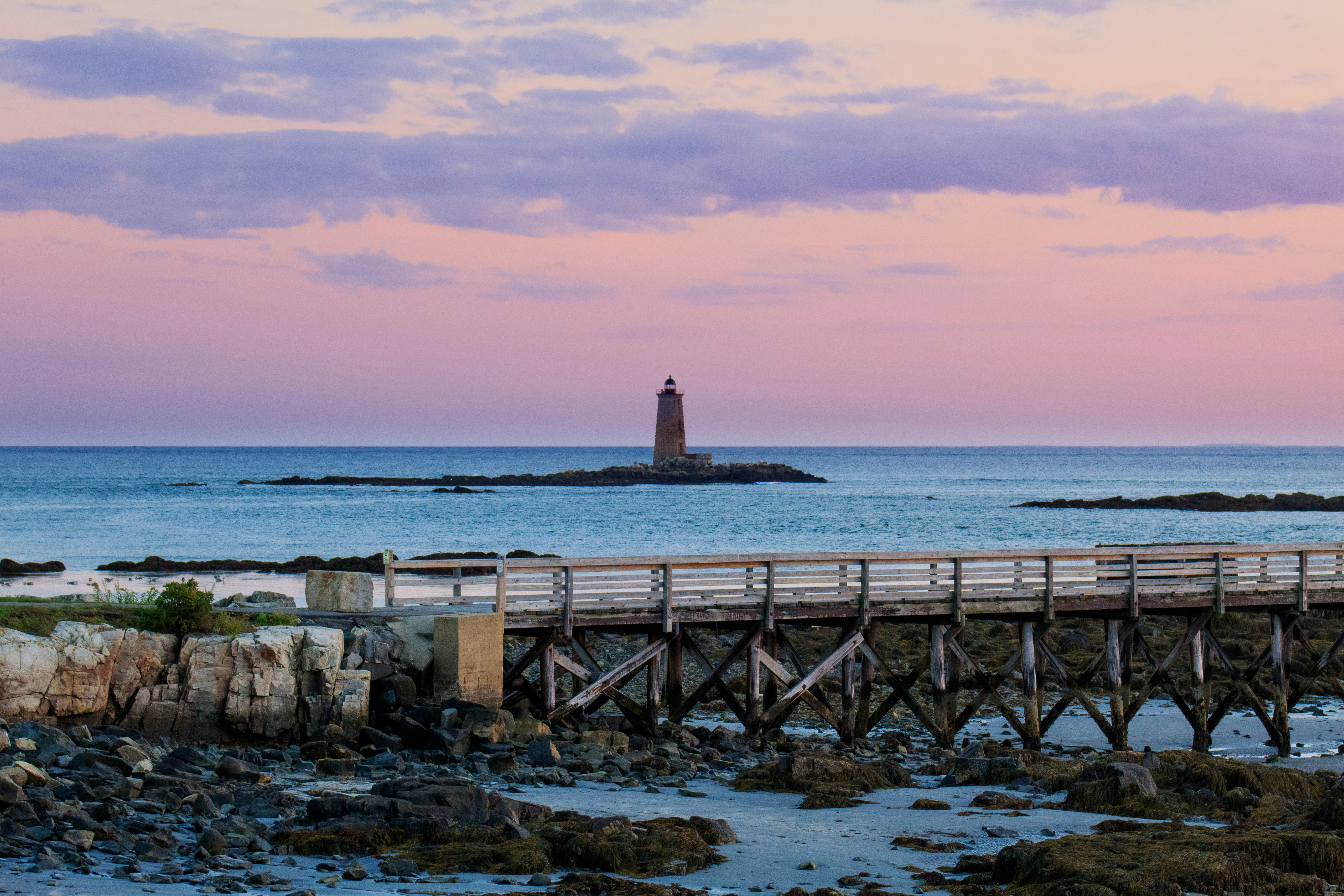 Lighthouse on a rocky islet beneath a pastel sunset sky with a wooden bridge in the foreground.
