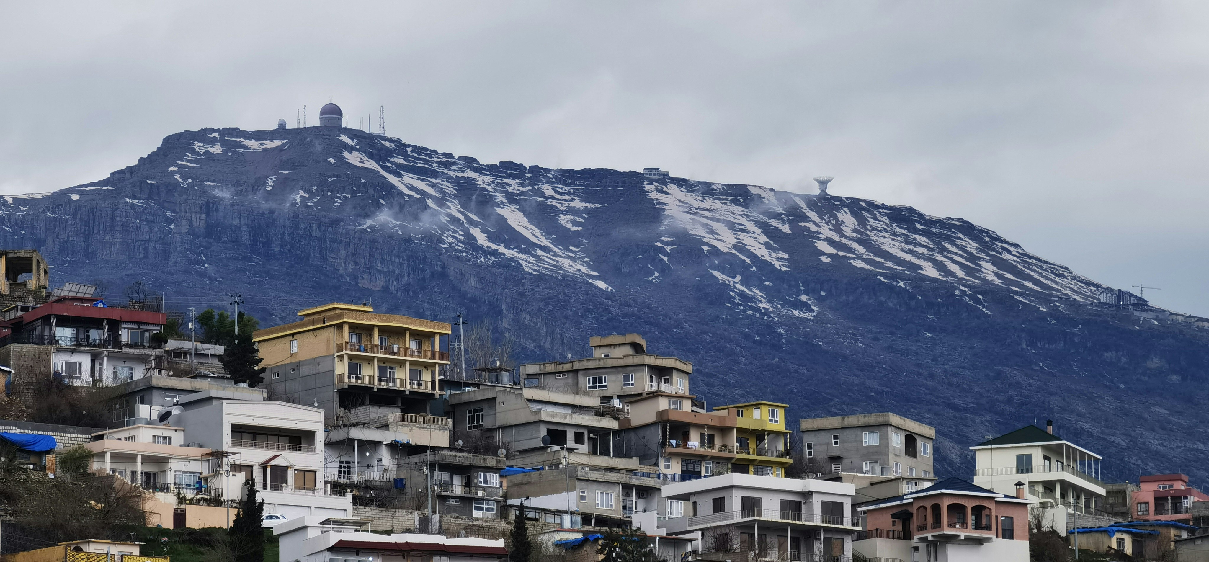 Clustered buildings rise against the backdrop of a snow-dusted mountain under a cloudy sky.