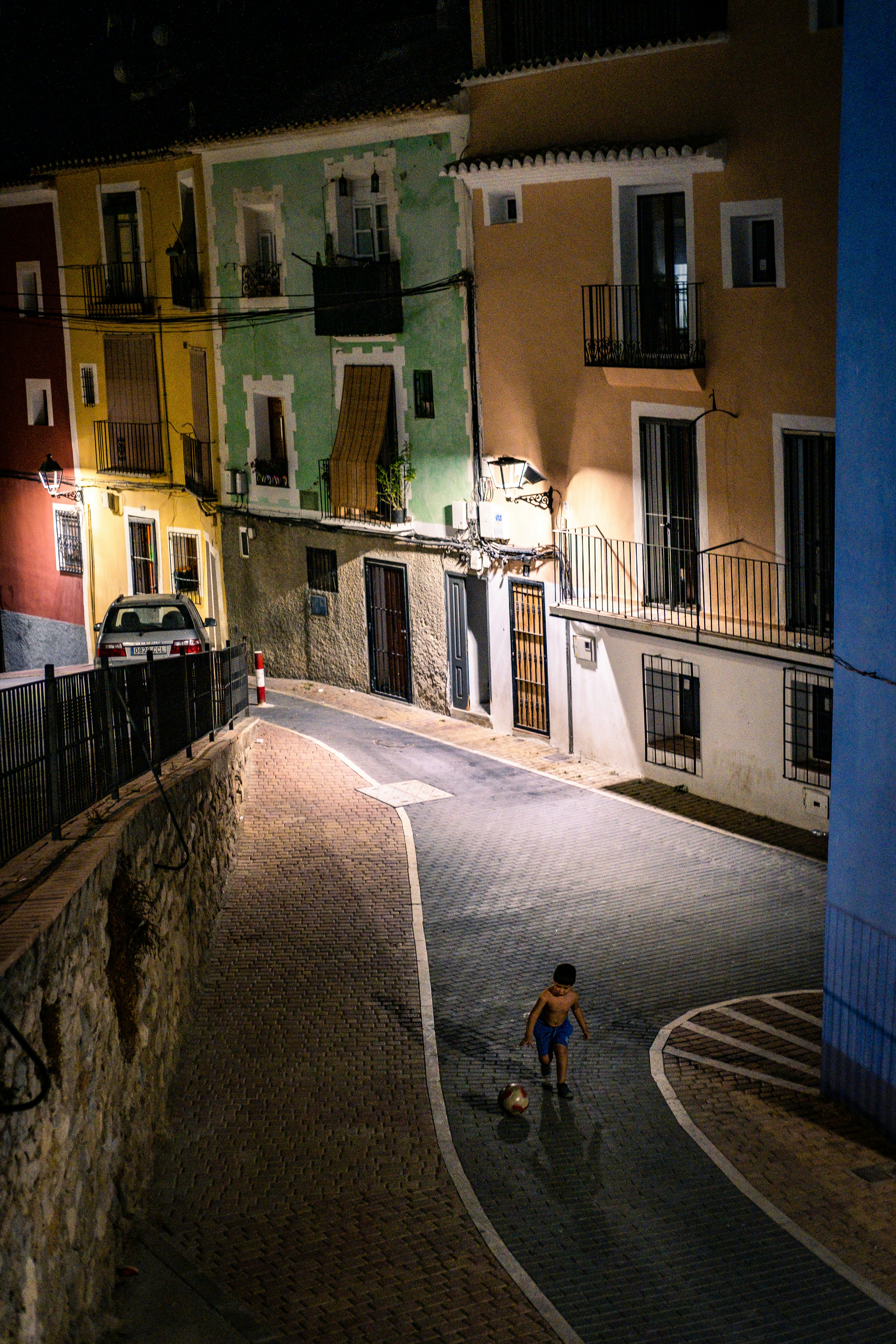 Moonlit Curve: A boy dribbles a ball through a winding, pastel-fronted street.