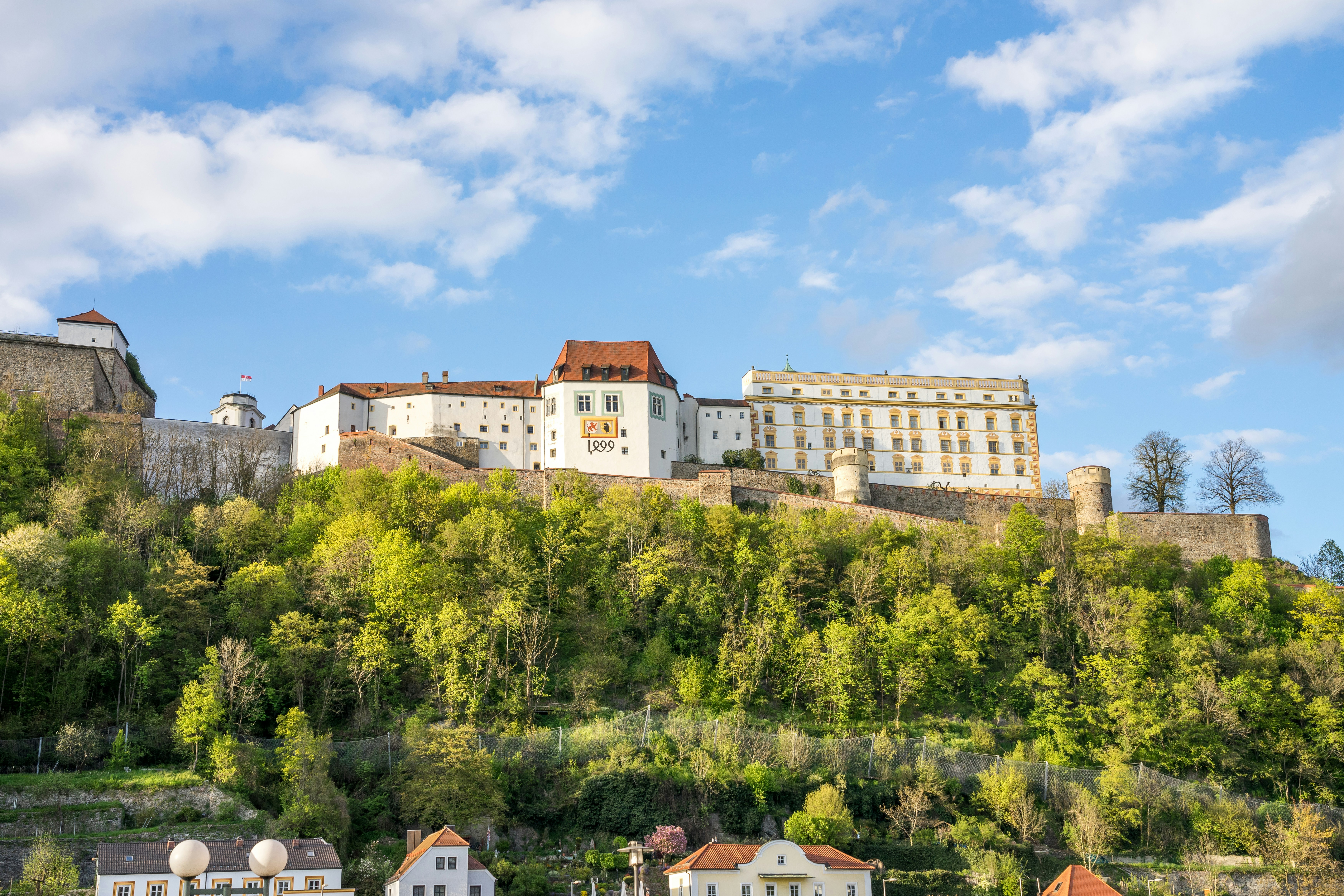 Castle perched atop a lush, tree-lined hill under a vibrant blue sky with scattered clouds.