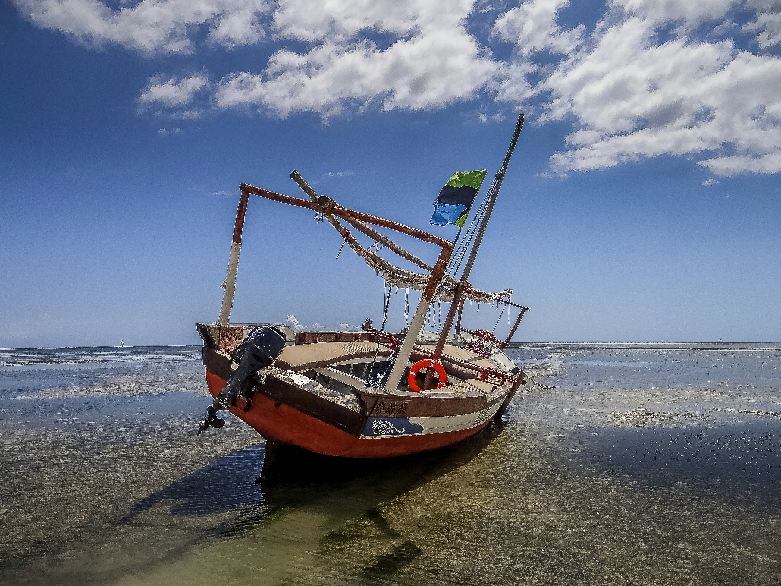 A small wooden boat sits in shallow, clear water with its red hull and weathered mast beneath a bright blue sky. A flag flutters at the mast, completing the maritime scene.