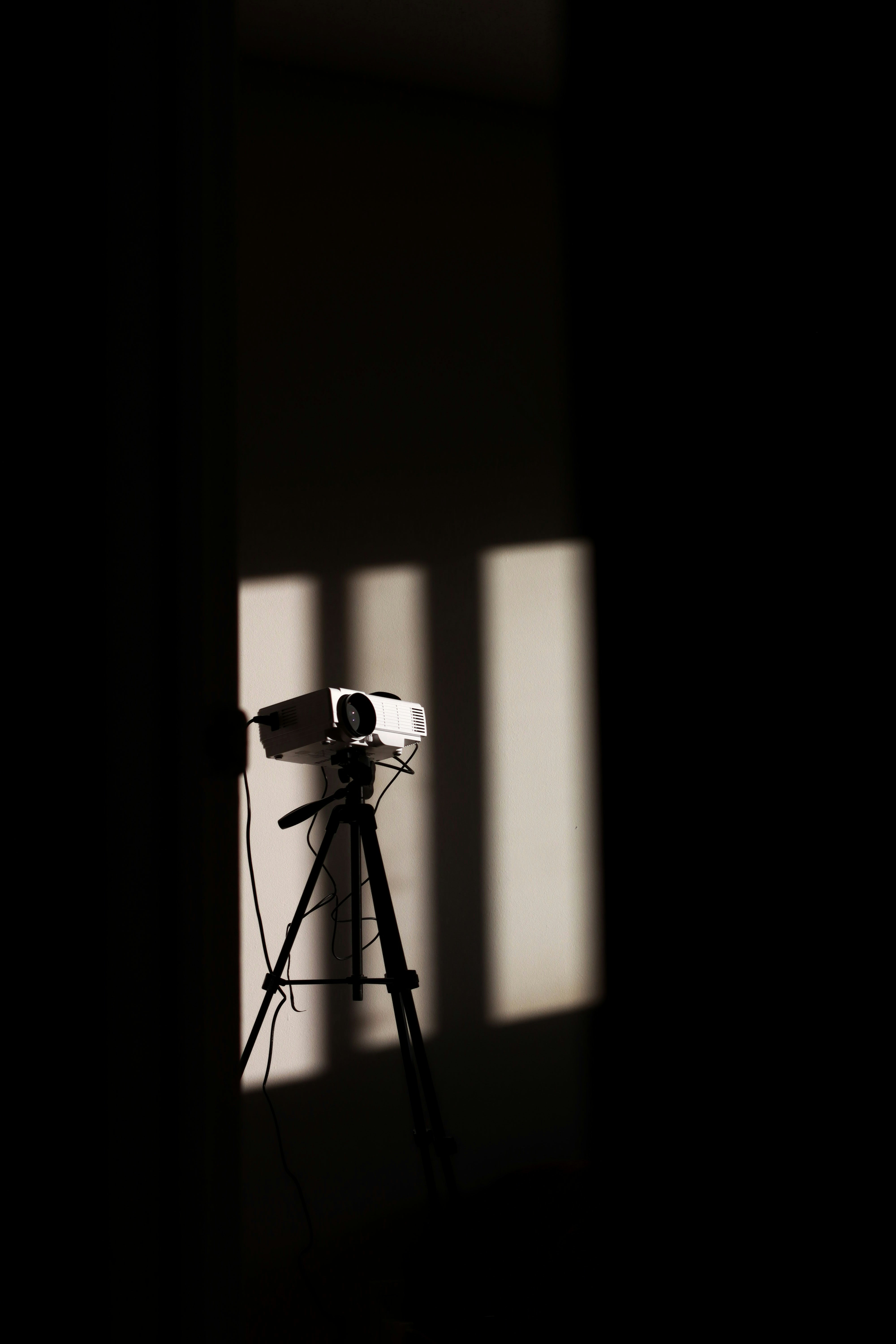 A lone projector stands on a tripod, casting shadows in a dimly lit room with light streaks from a nearby window.