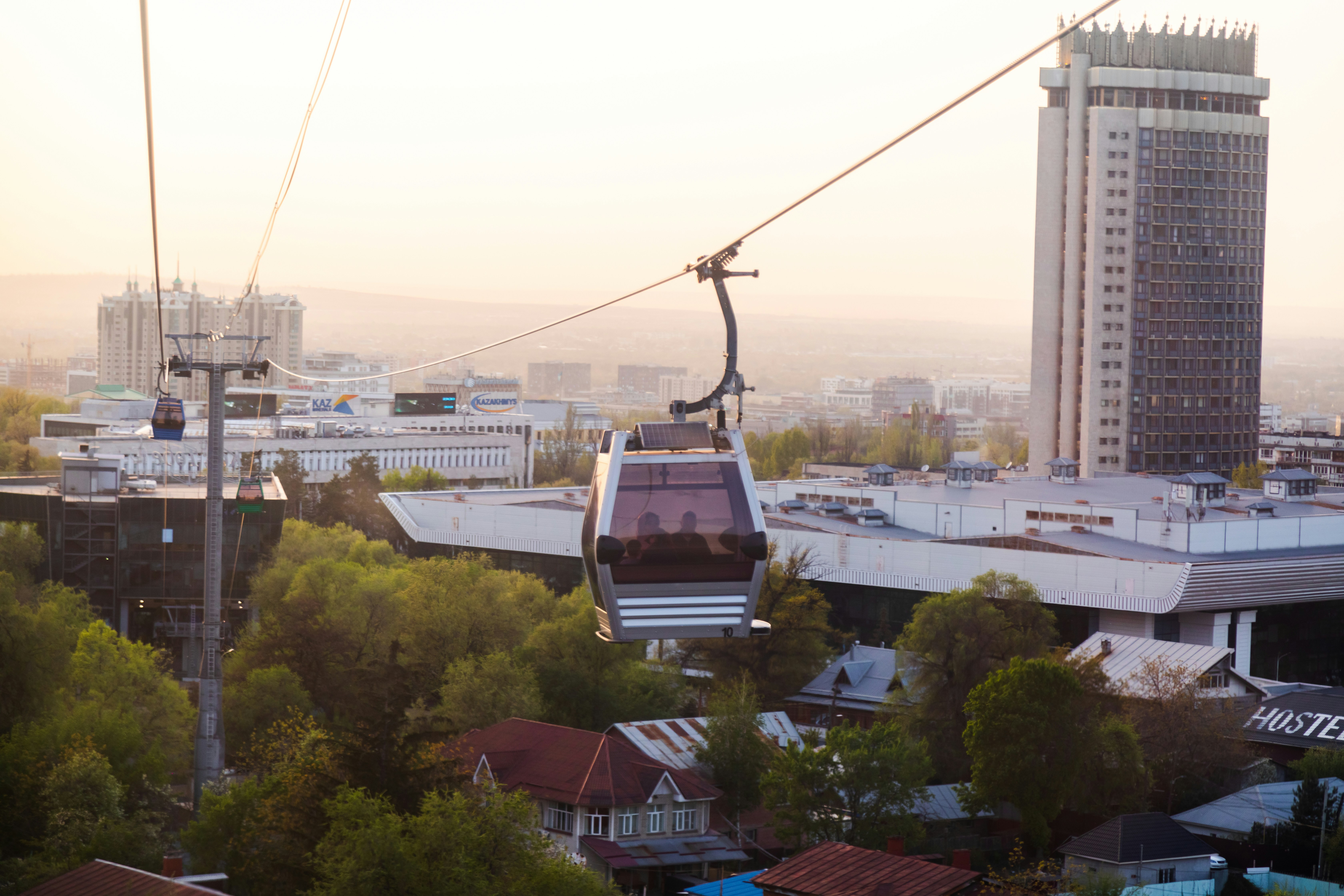 Cable car gliding above a city landscape with a tall building and trees at dusk.