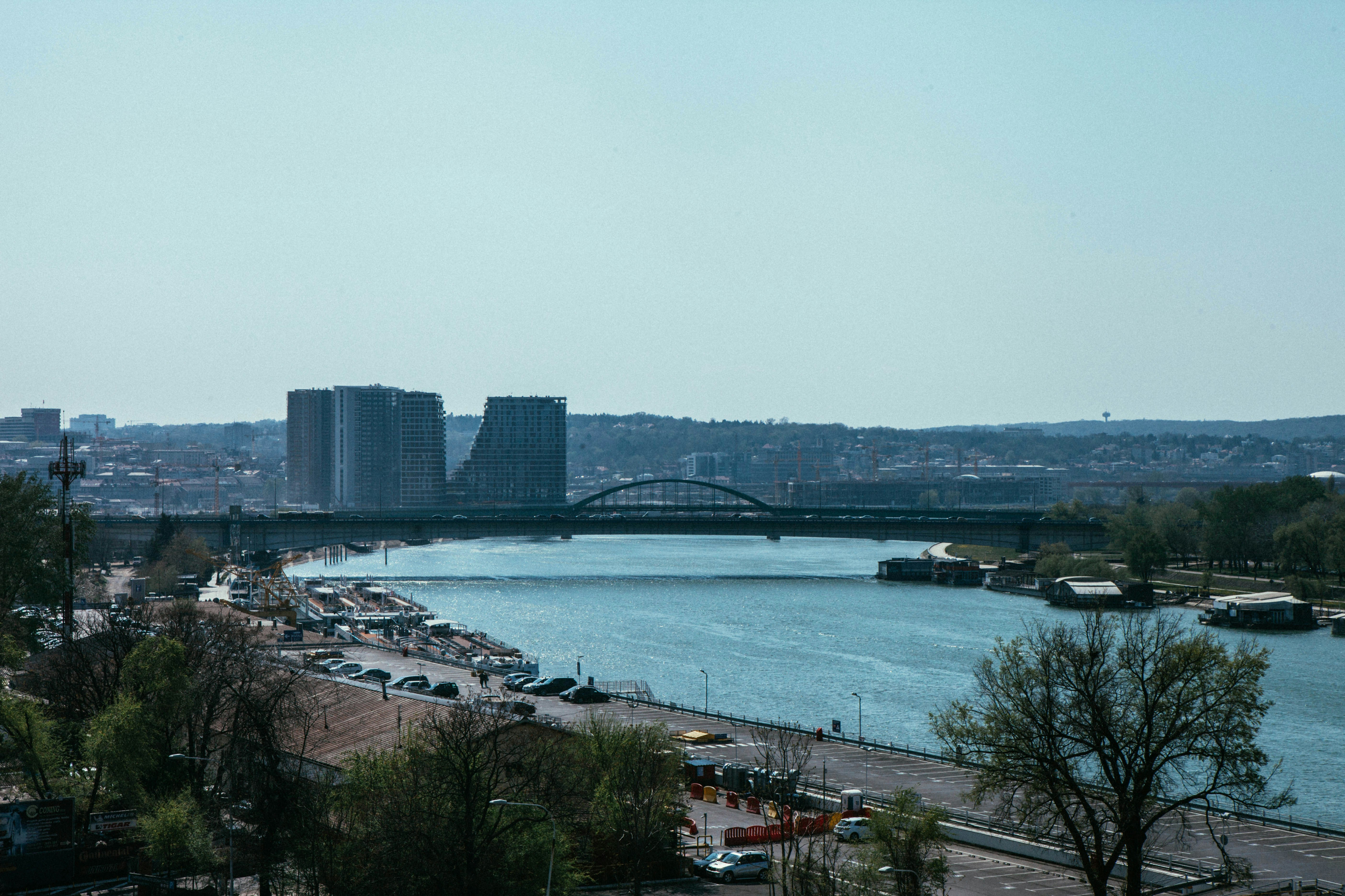 A calm river flanked by a busy promenade and a distant bridge, set against a cityscape under a clear sky.