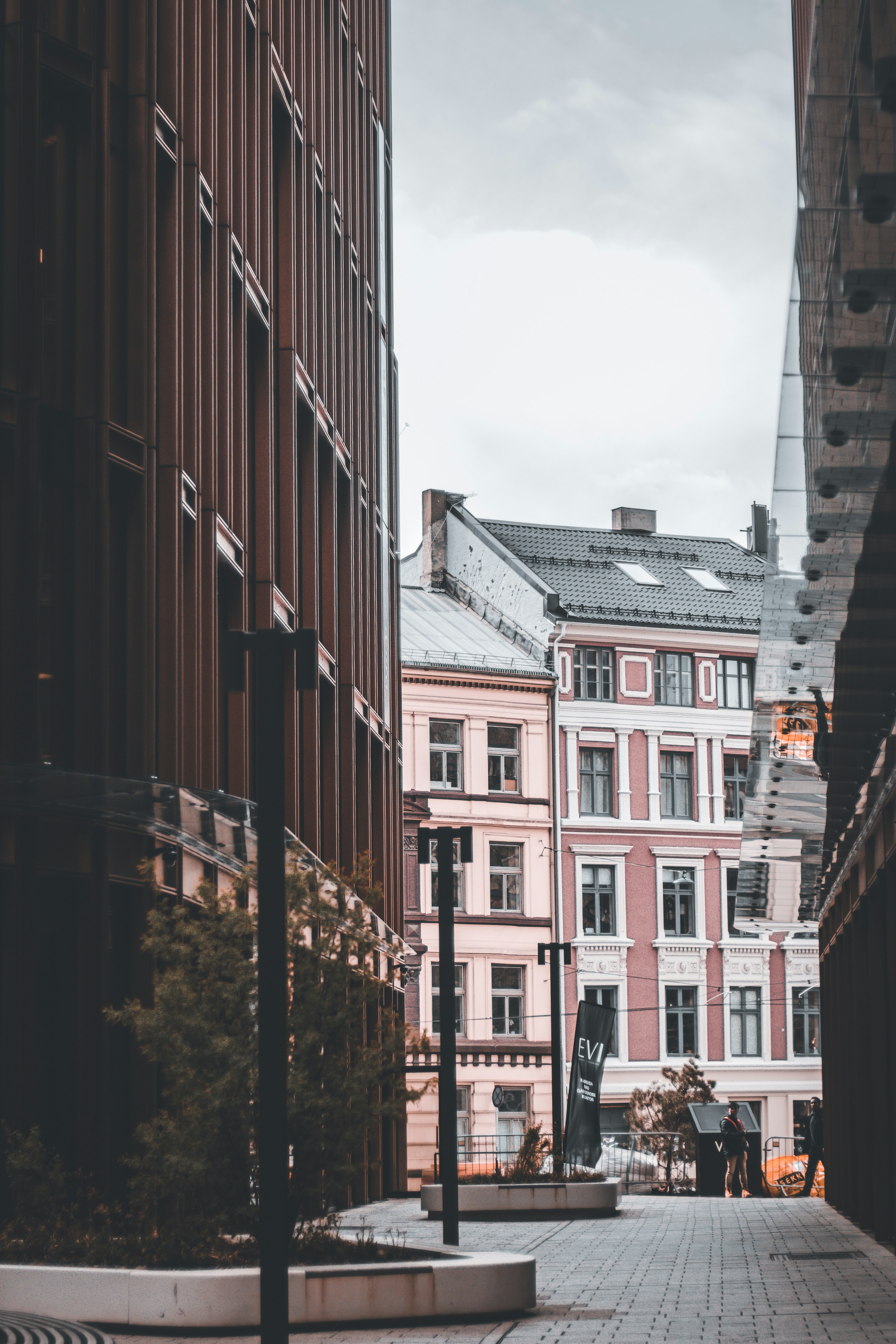 Narrow alleyway framed by tall buildings, revealing a glimpse of colorful architecture in the background. A blend of modern and classic design elements creates an intriguing urban scene.