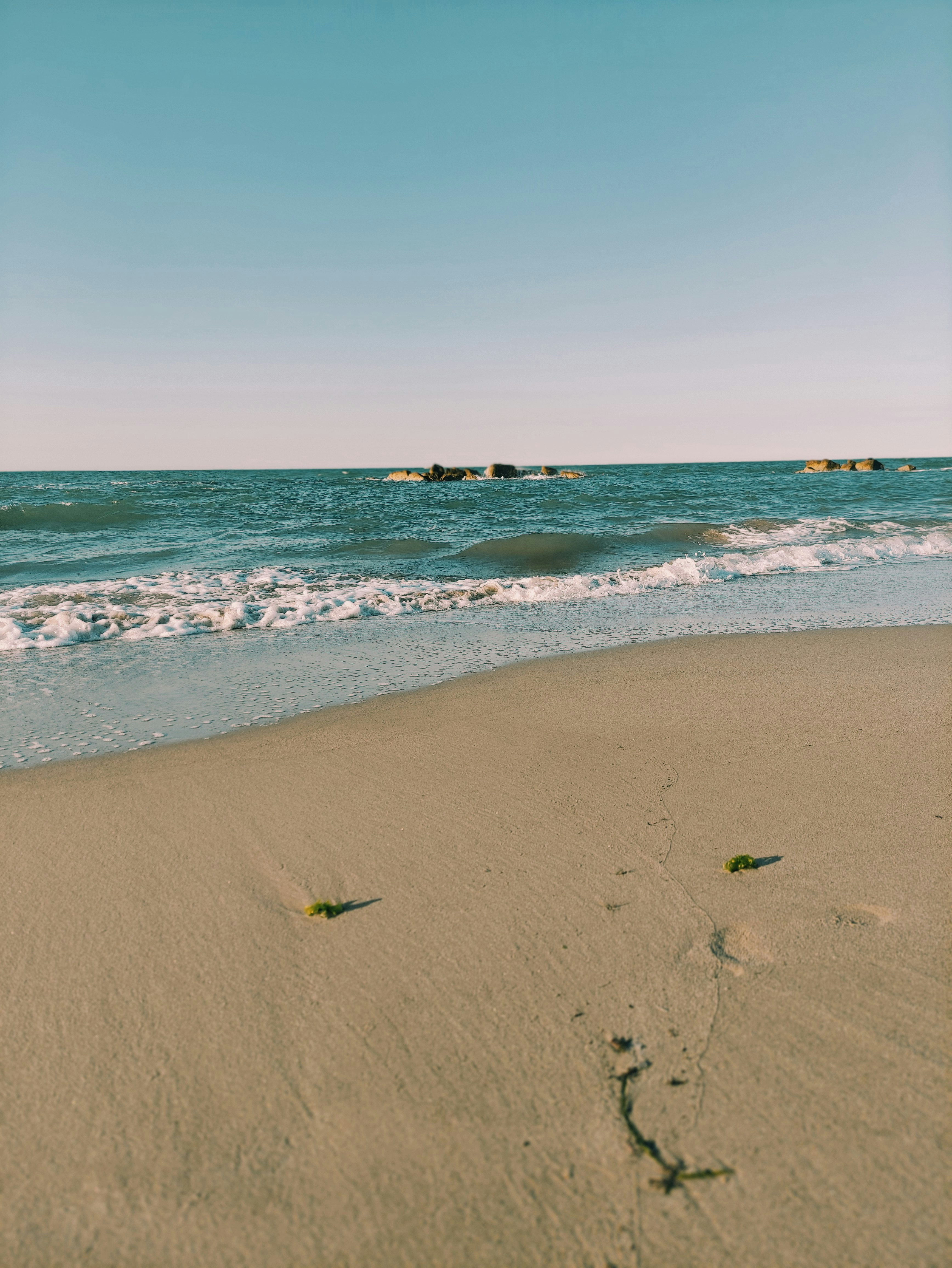 Sunlit beach with gentle waves and distant rock outcrops on the horizon under a gradient blue sky.